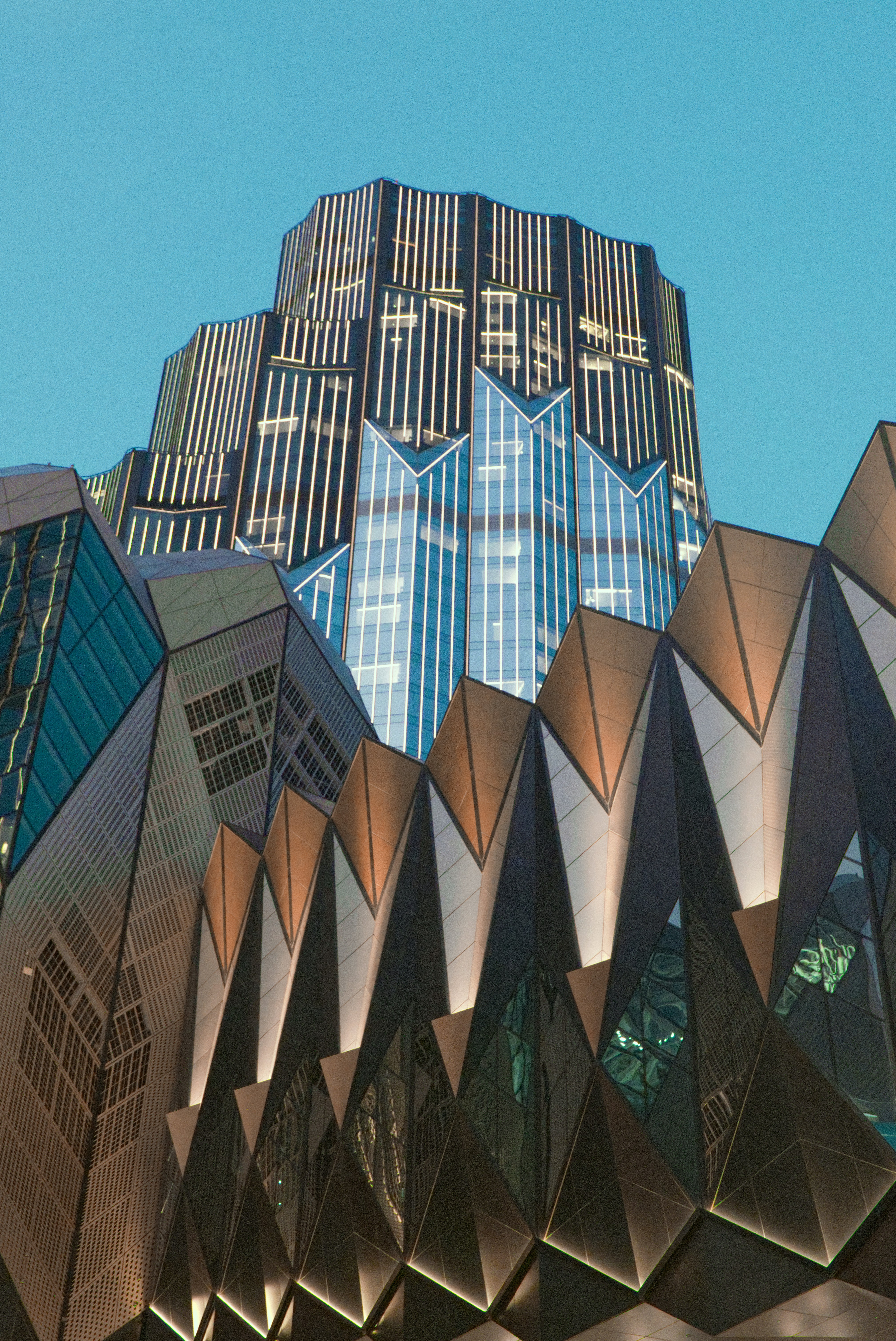 Geometric skyscraper facades rise with angular copper-toned panels against a clear blue sky, their sharp peaks mirrored by glassy windows.