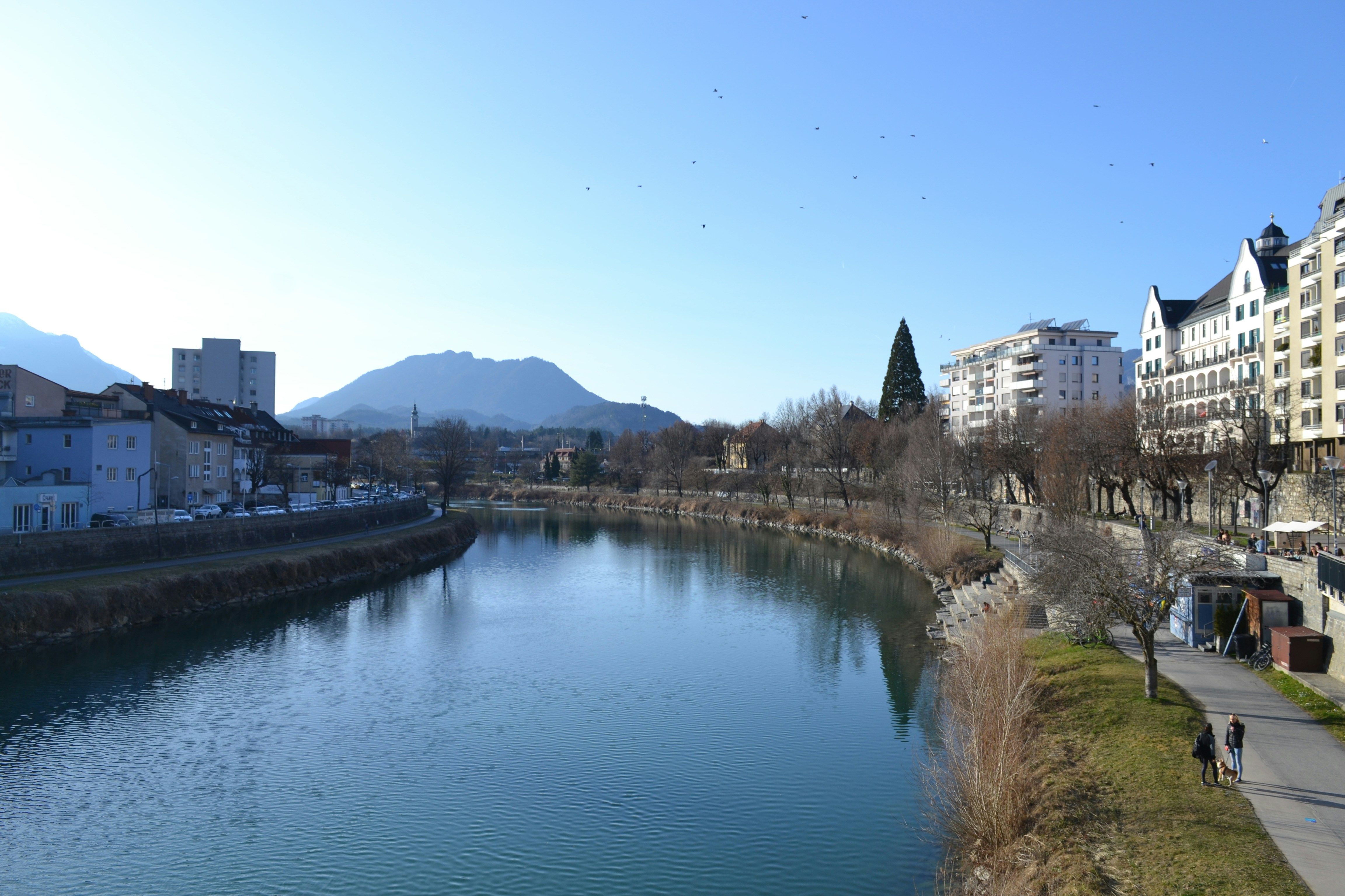 Riverwalk with Alpine Backdrop: Sunlit Cityscape Along a Calm Canal