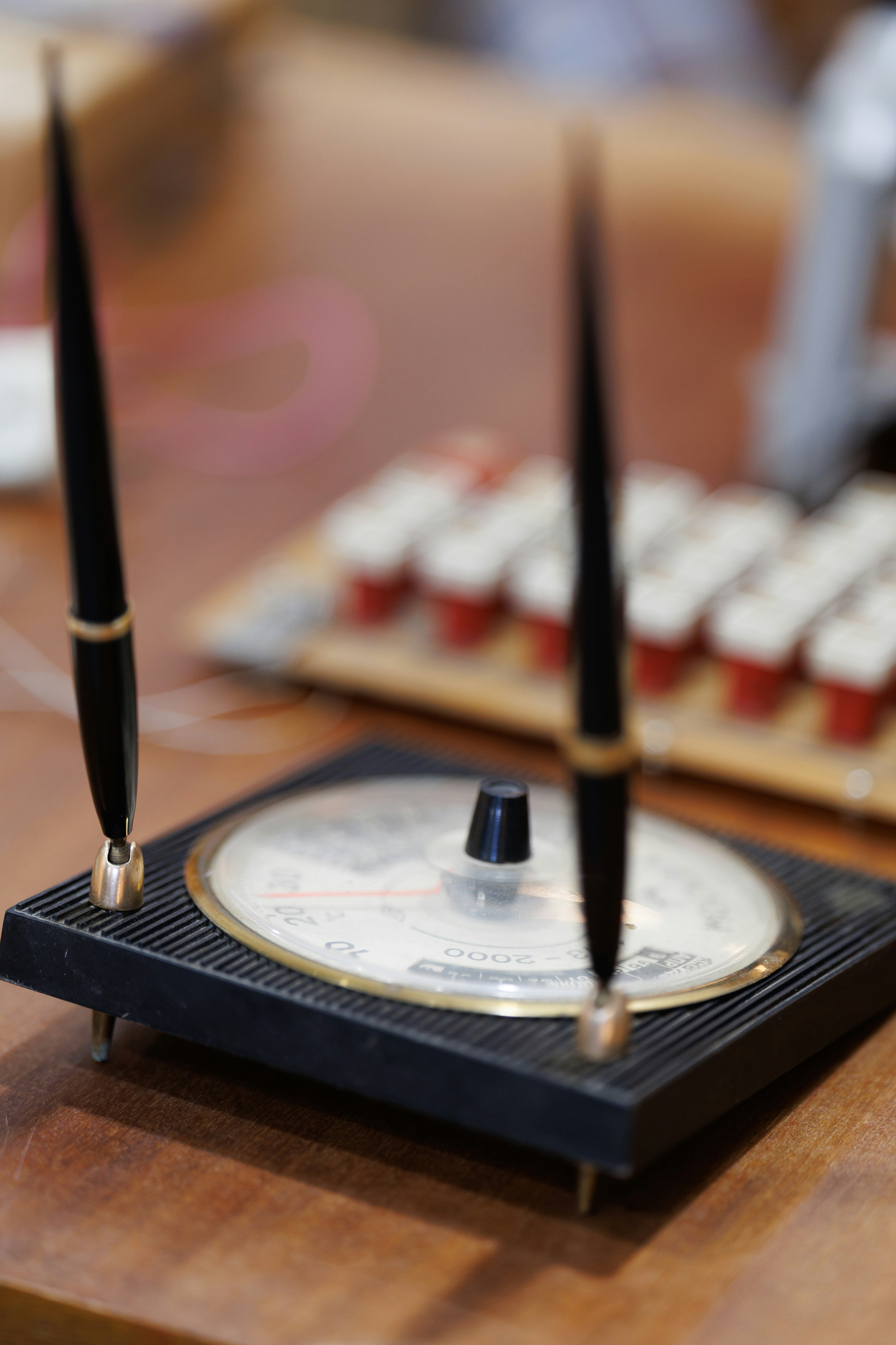 Close-up of a vintage dial timer with two slender tuning forks facing it, resting on a wooden surface; shallow depth of field blurs the background with hints of a manufacturing or plotting setup.