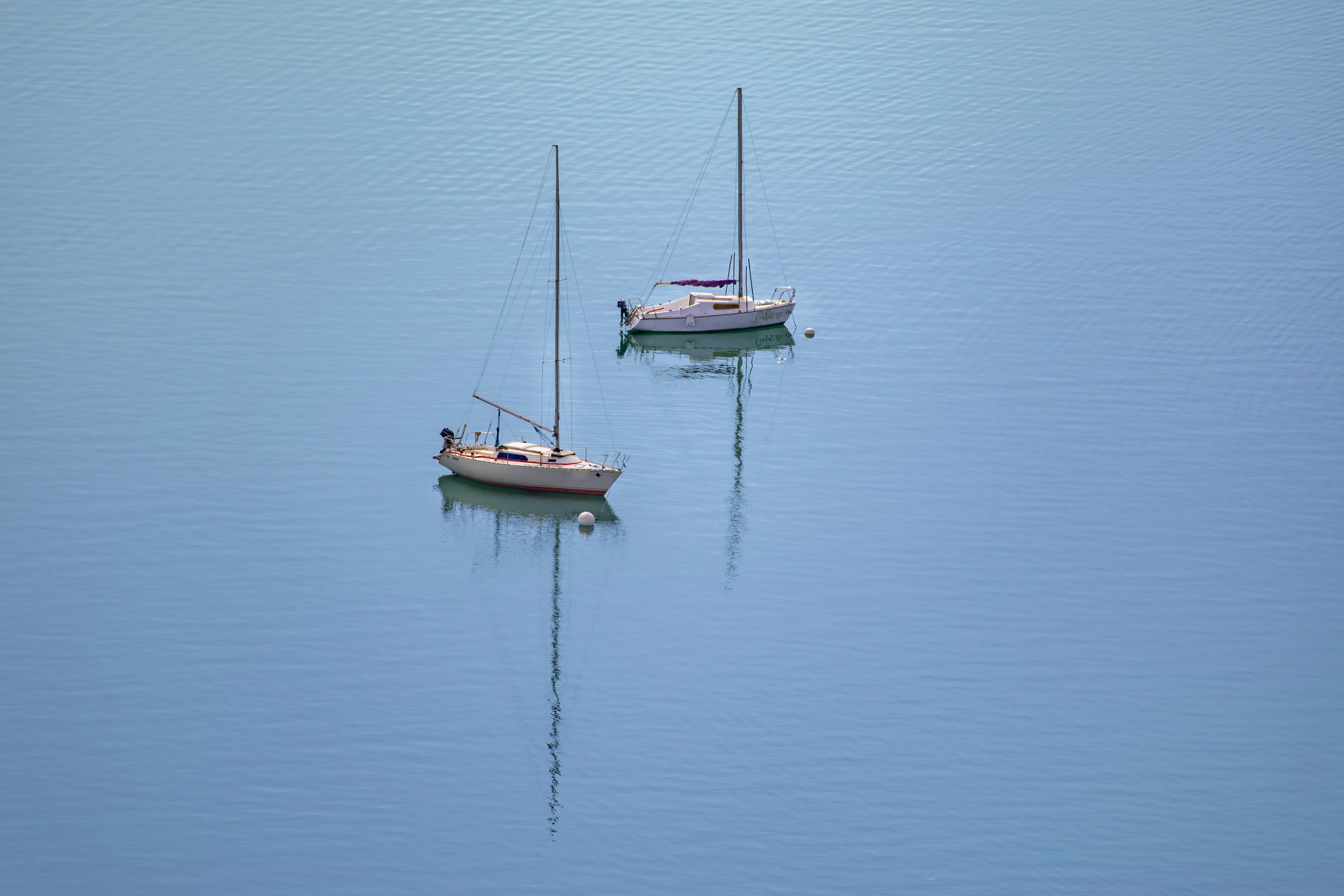 Two sailboats float on a calm blue sea with their masts rising, creating mirrored reflections beneath. A minimal, serene coastal scene captured from above.