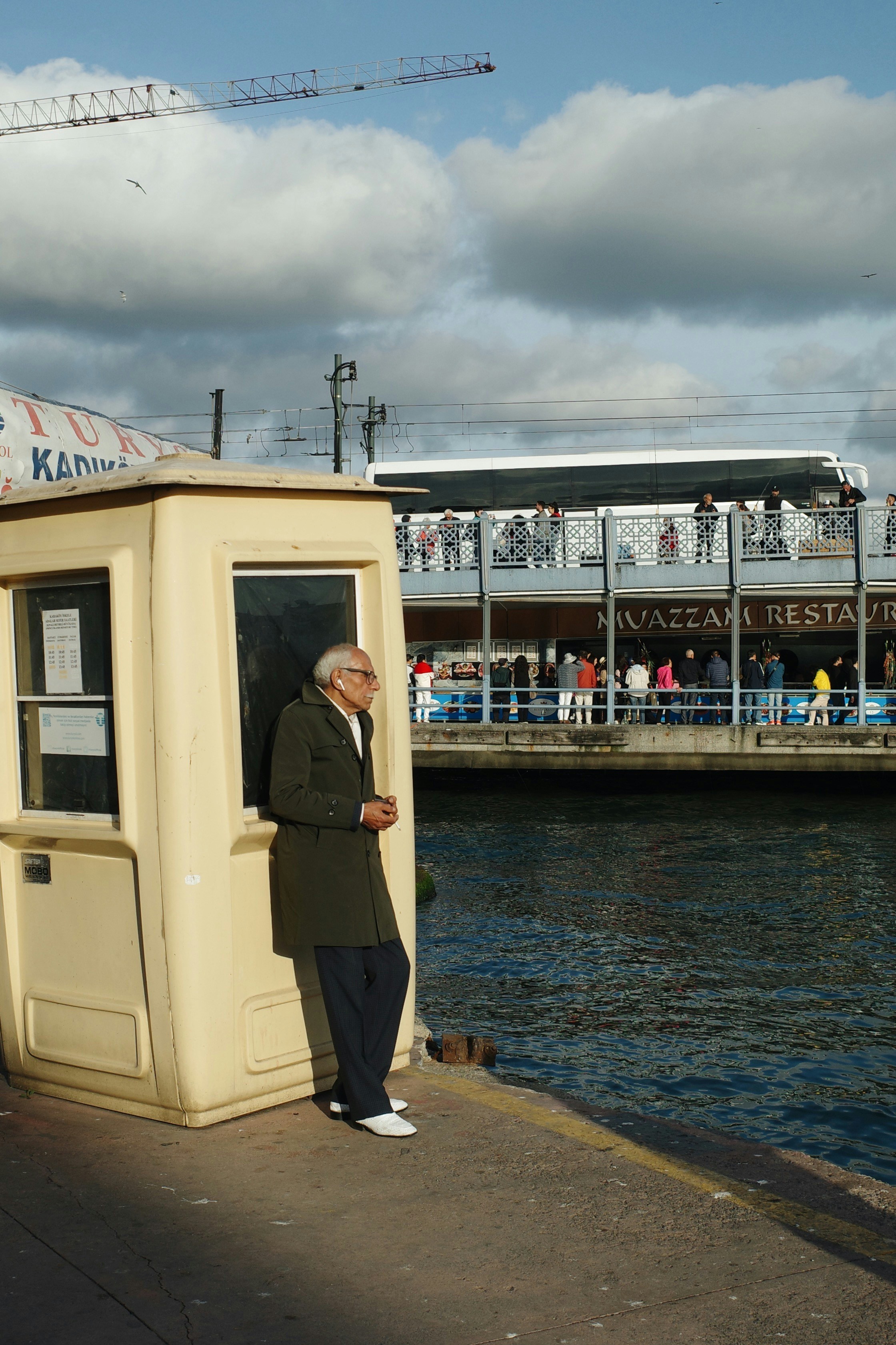 Man in a Coat Leaning by a Dockside Booth as a Ferry Sails by