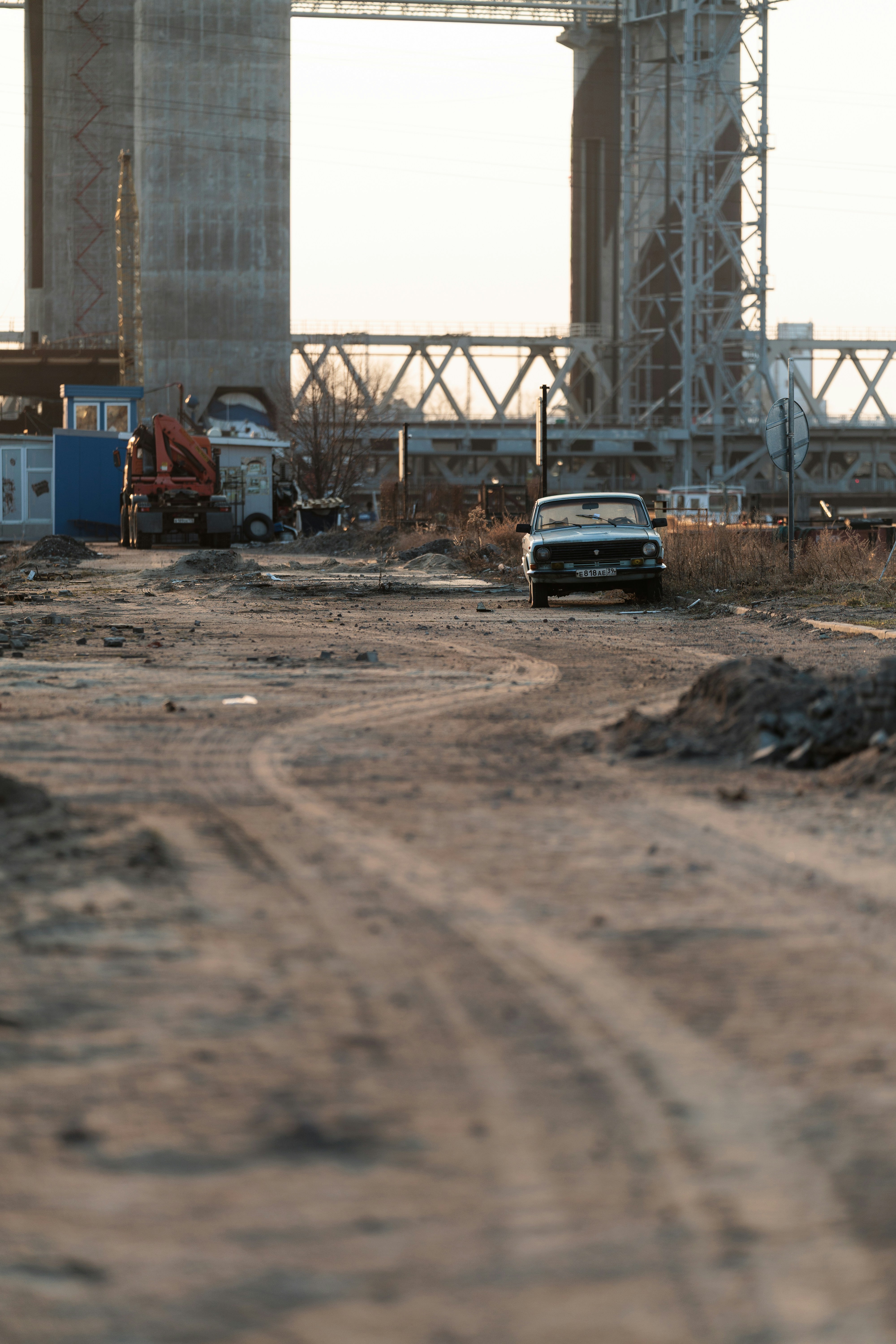 Rusty sedan sits amid a desolate construction site, with industrial structures looming in the background and a dirt road leading the eye toward the vehicle.