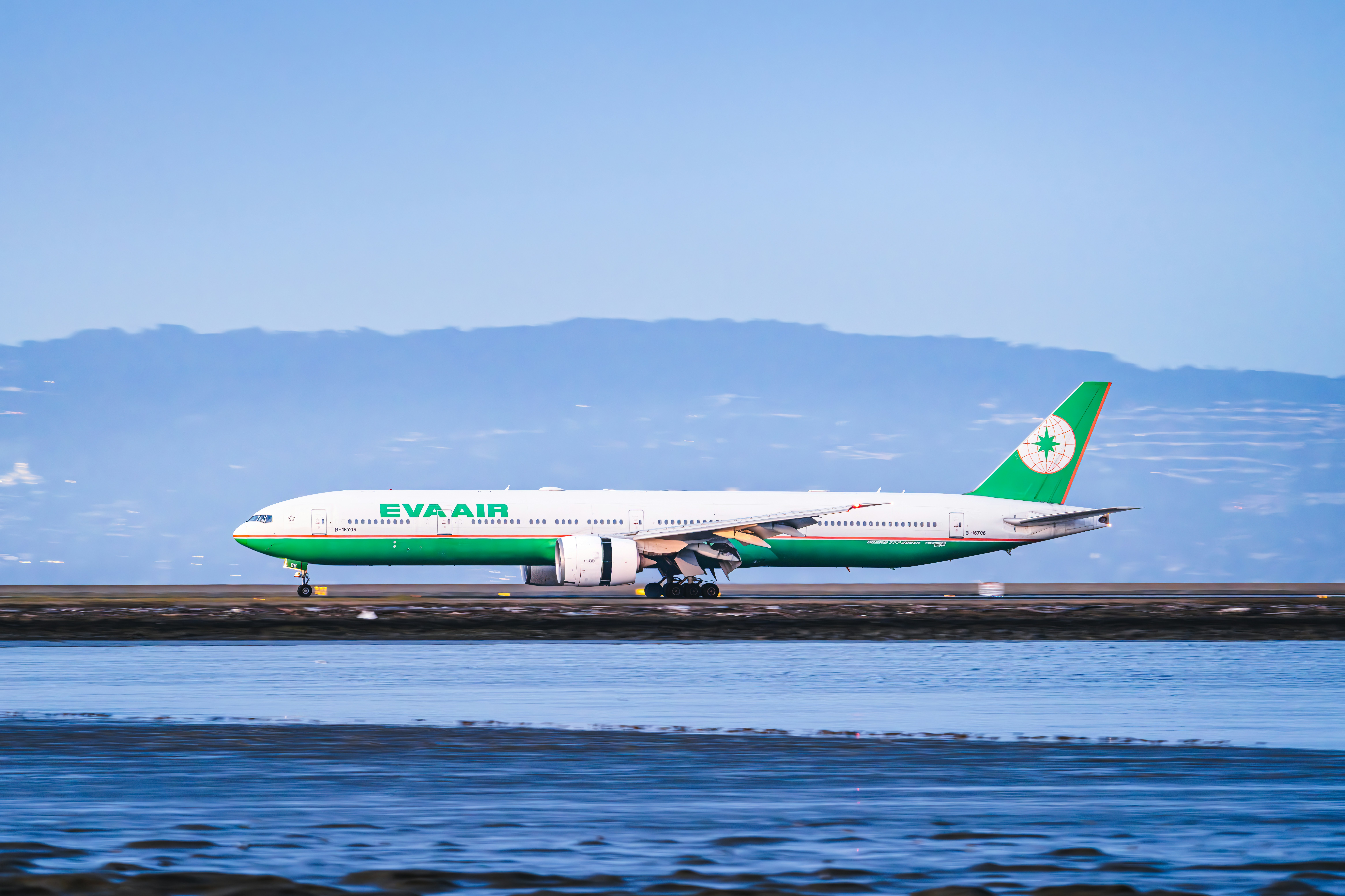 Eva Air 777 on a distant runway, calm water foreground, blue sky