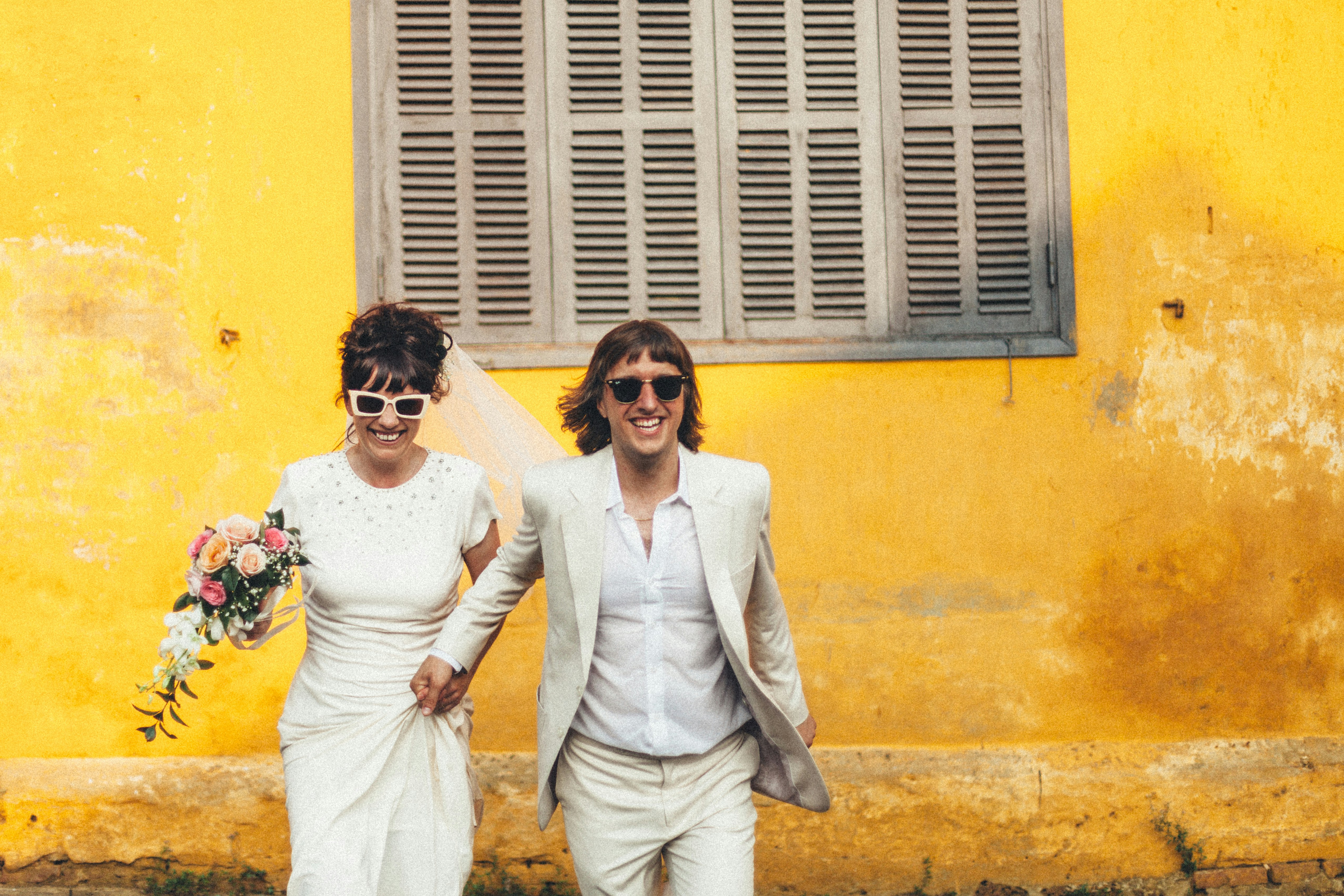 Bridal duo strides through sunlit yellow facade, laughter in the air