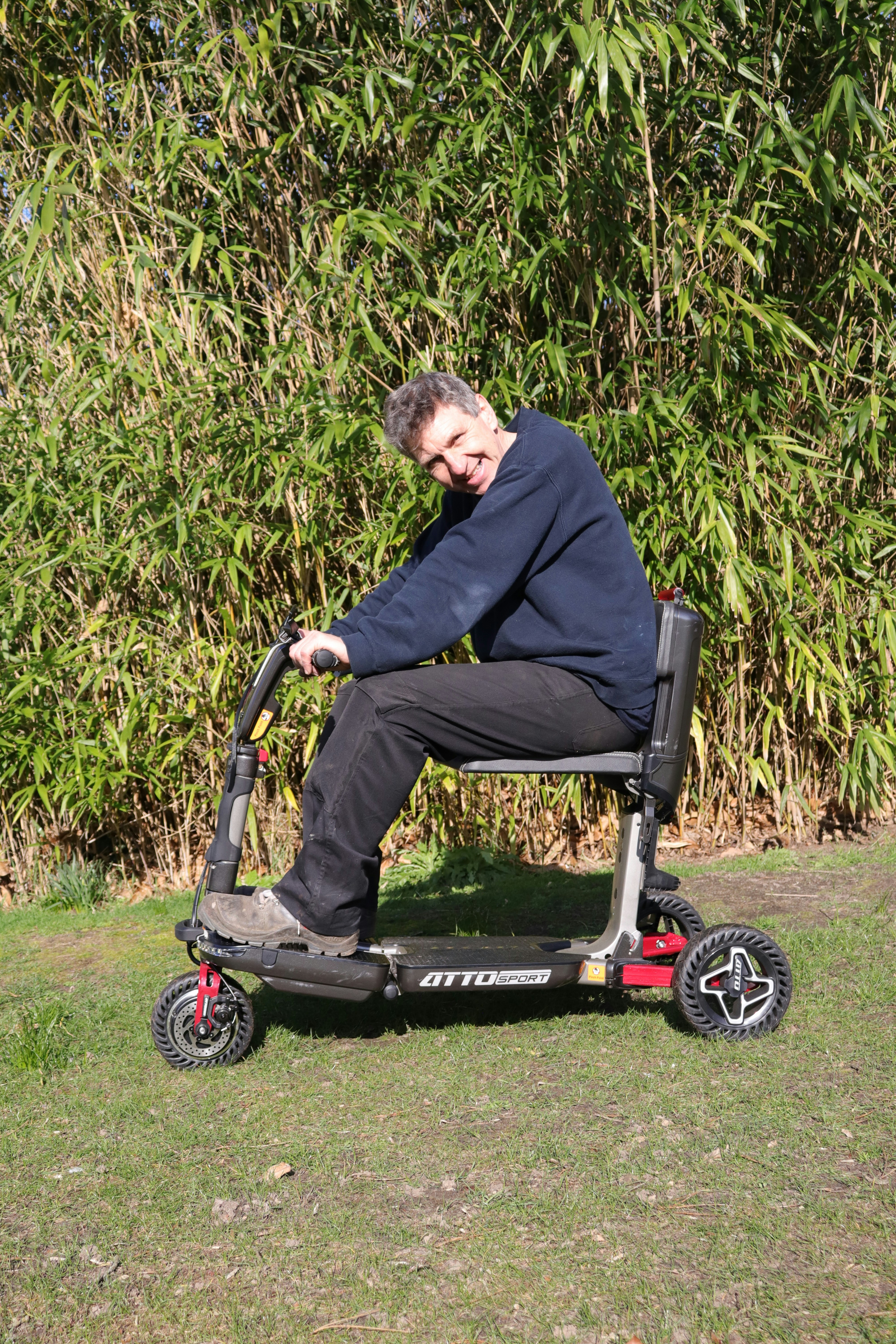 Rider in a Quiet Park: A man in a navy sweater sits on a low-slung electric scooter against a backdrop of tall bamboo and greenery