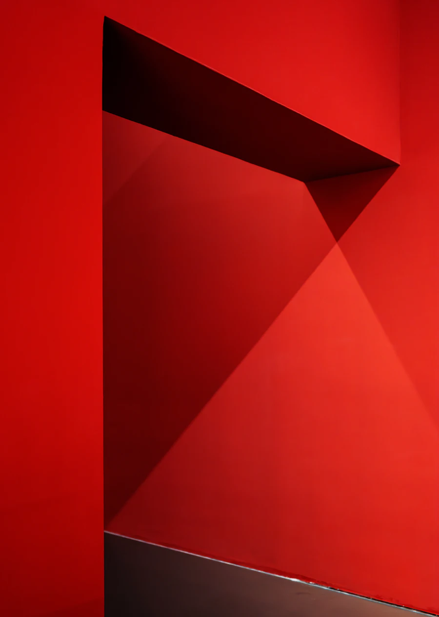 Red abstract interior with bold geometric planes converging at a diagonal junction; the composition emphasizes strong color blocks and shadow play. (Note: abstract geometric composition, not a portrait or logo).