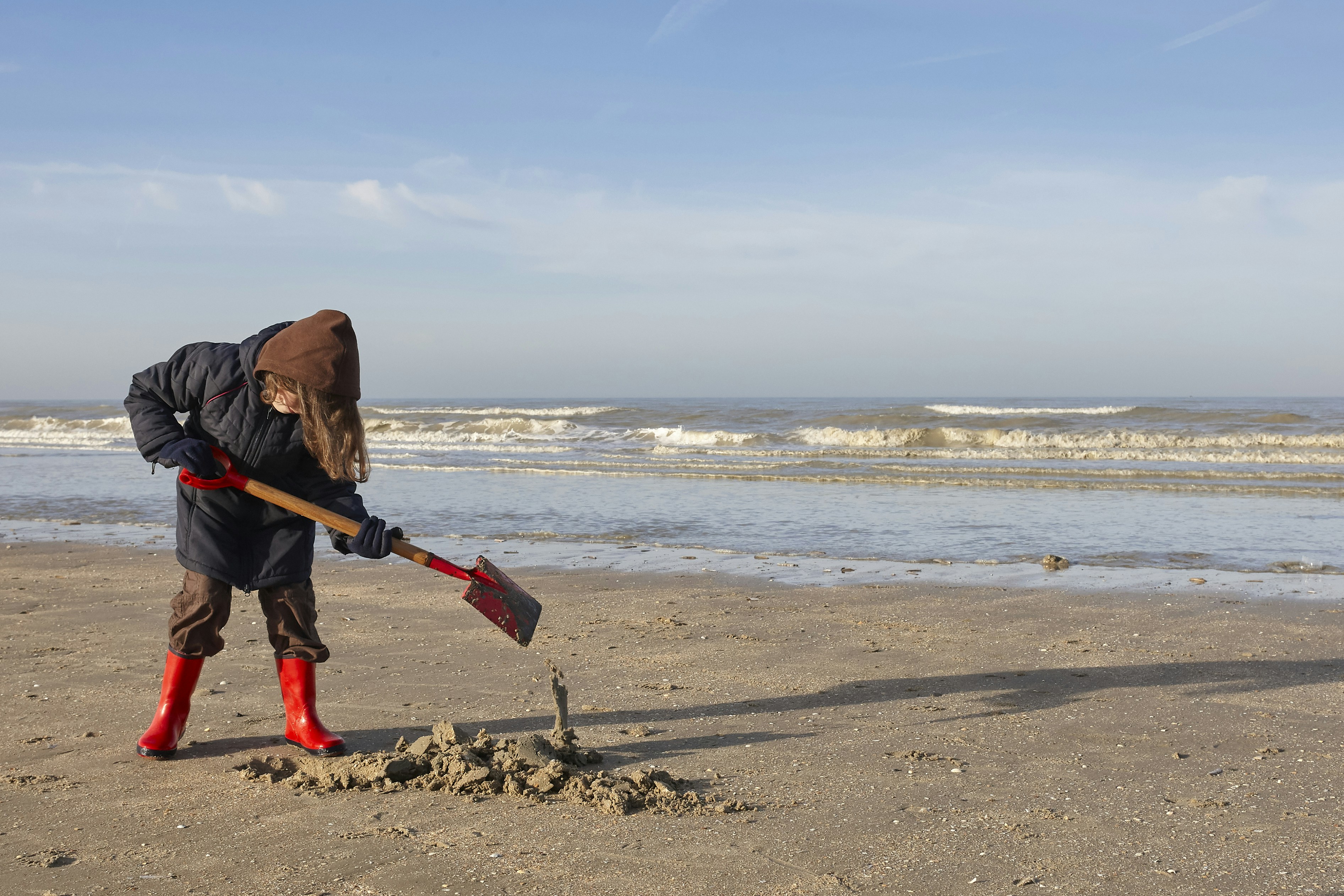 A child in red boots enthusiastically digs sand with a shovel on a sunny beach, with ocean waves gently breaking in the background under a clear blue sky.