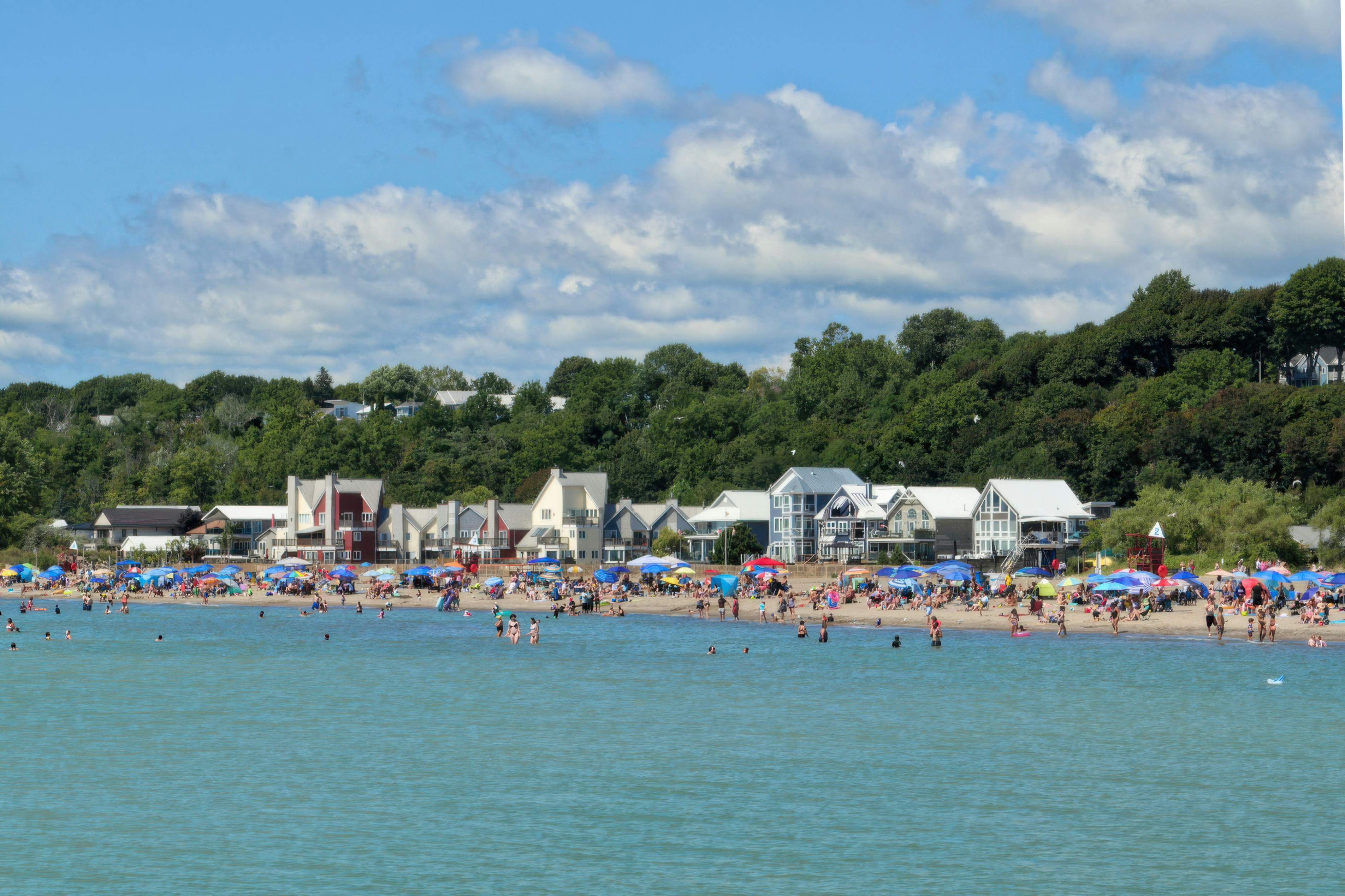 Crowded beach with buildings and trees under a cloudy sky.
