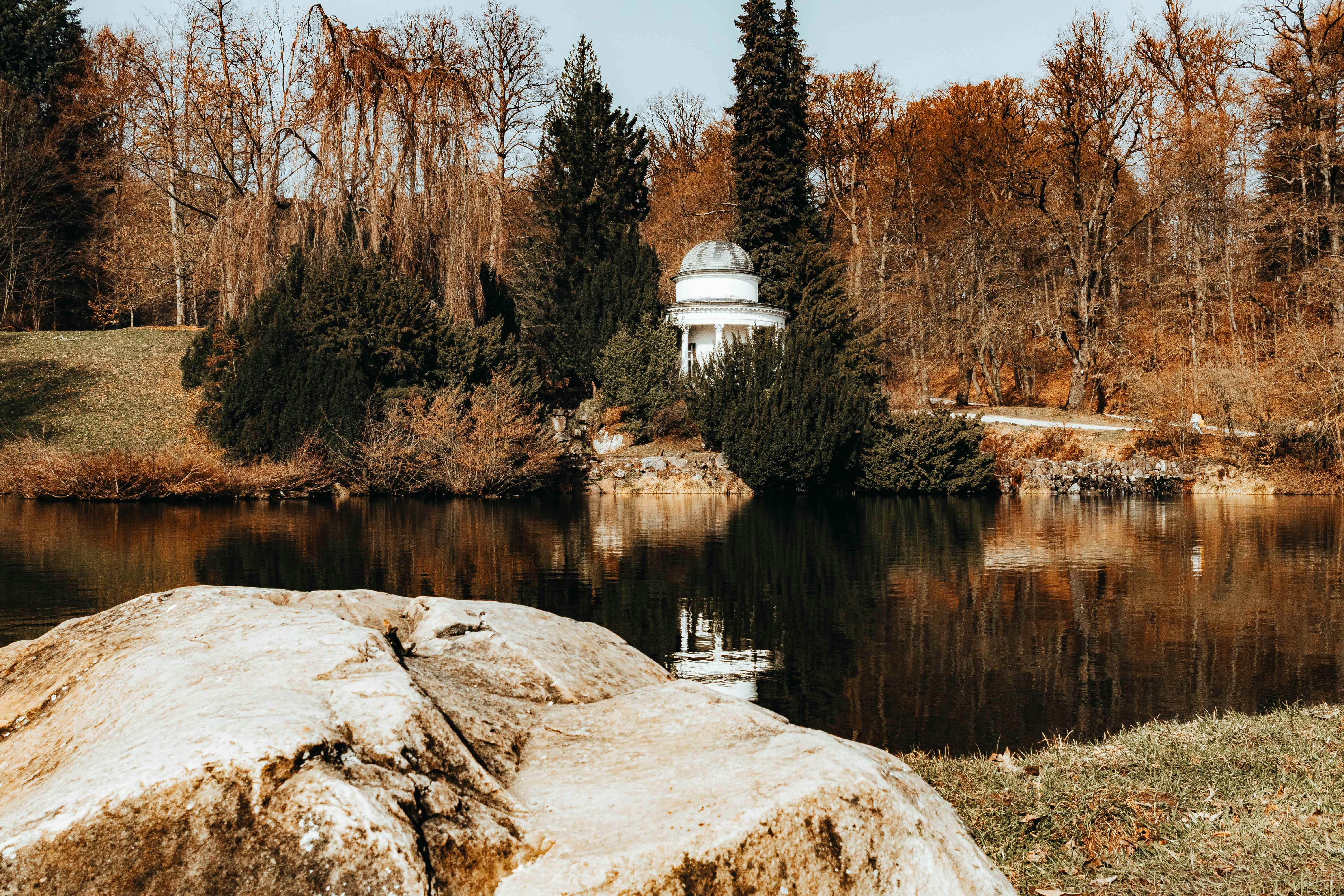 Stone foreground, tranquil lake, and white pavilion under autumn trees