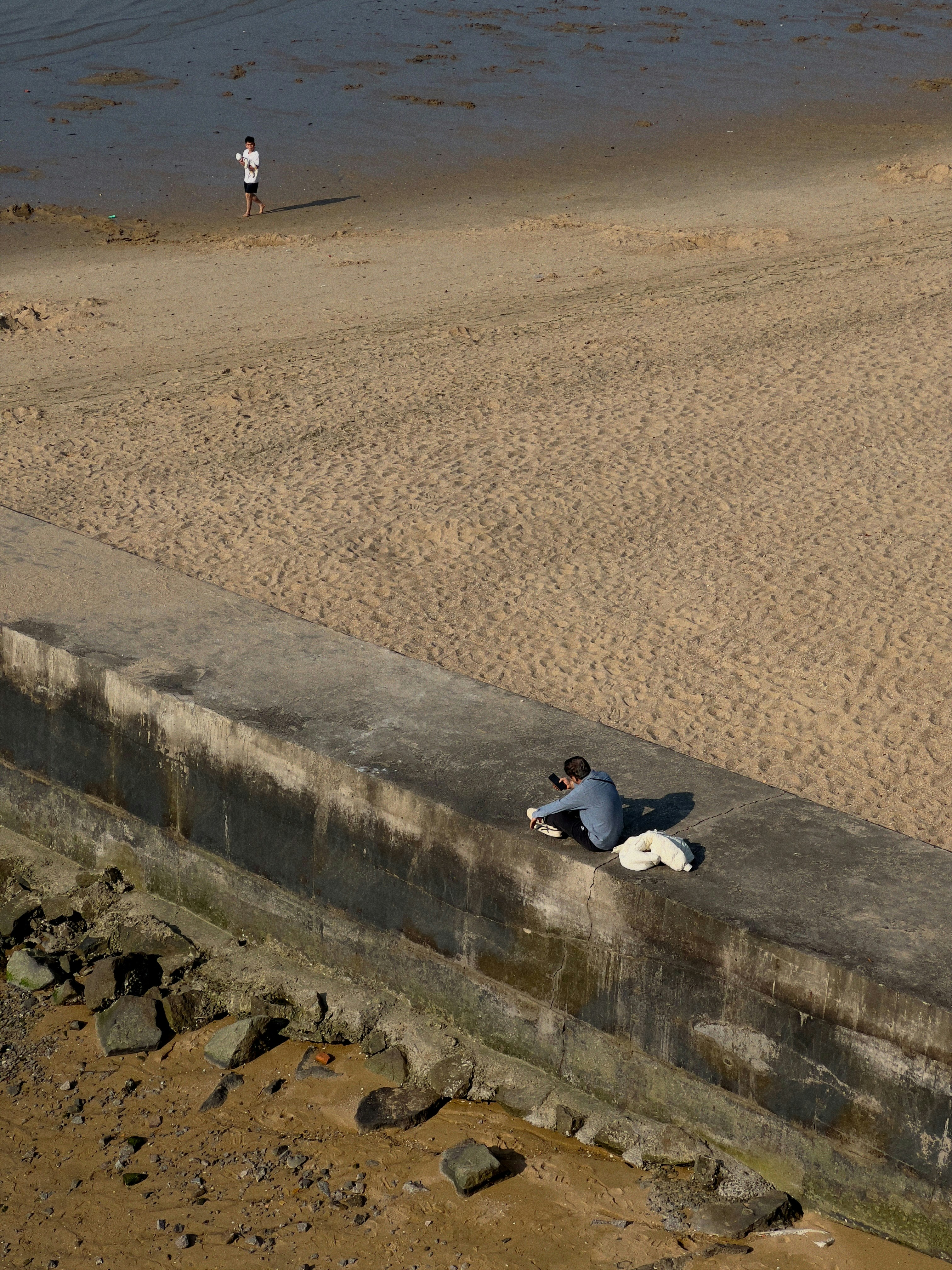 Two figures by the sea: a man on the shore and a person resting on a concrete ledge