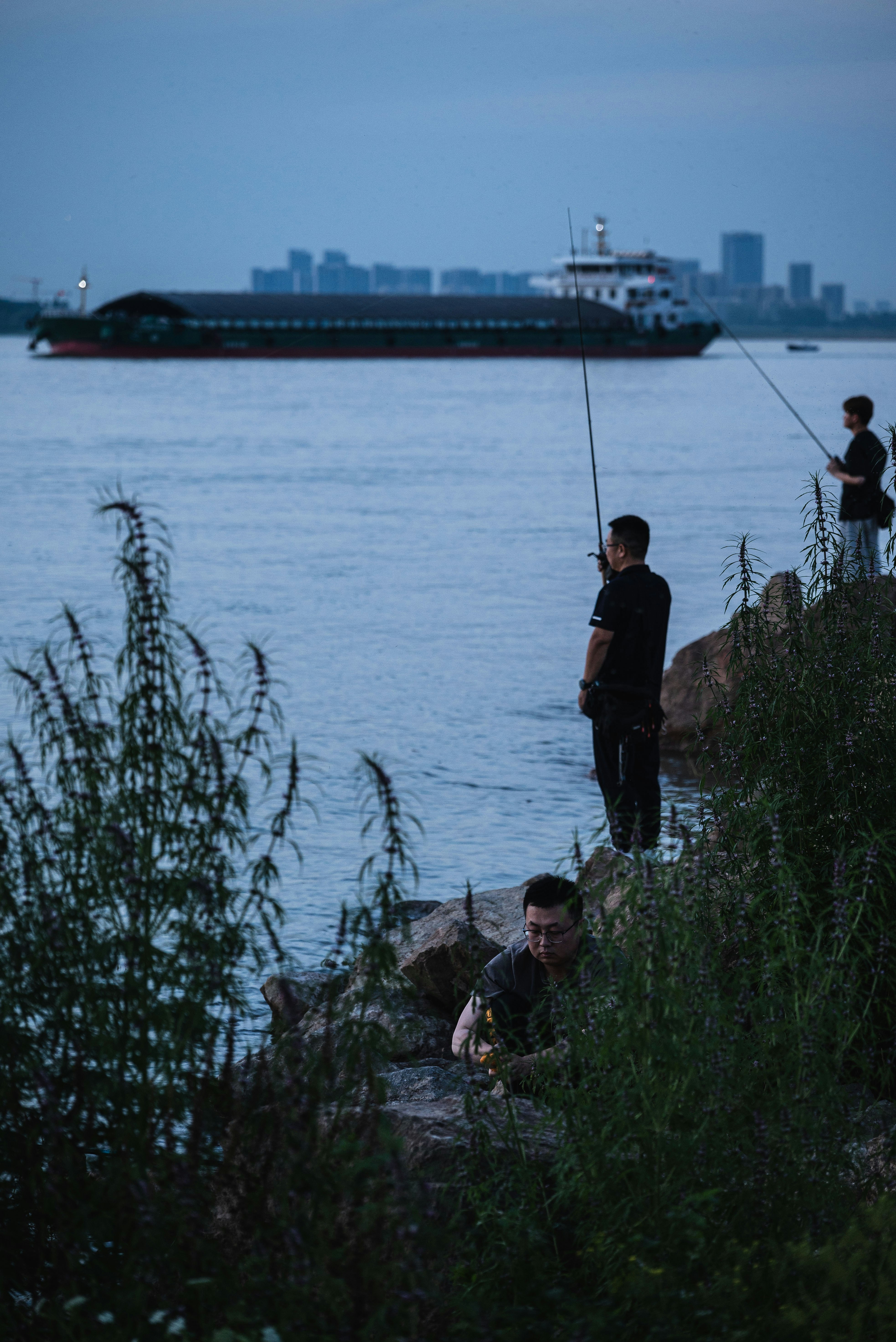 A Quiet Riverbank: Fishermen at Dusk with Wakeful Cargo Ship