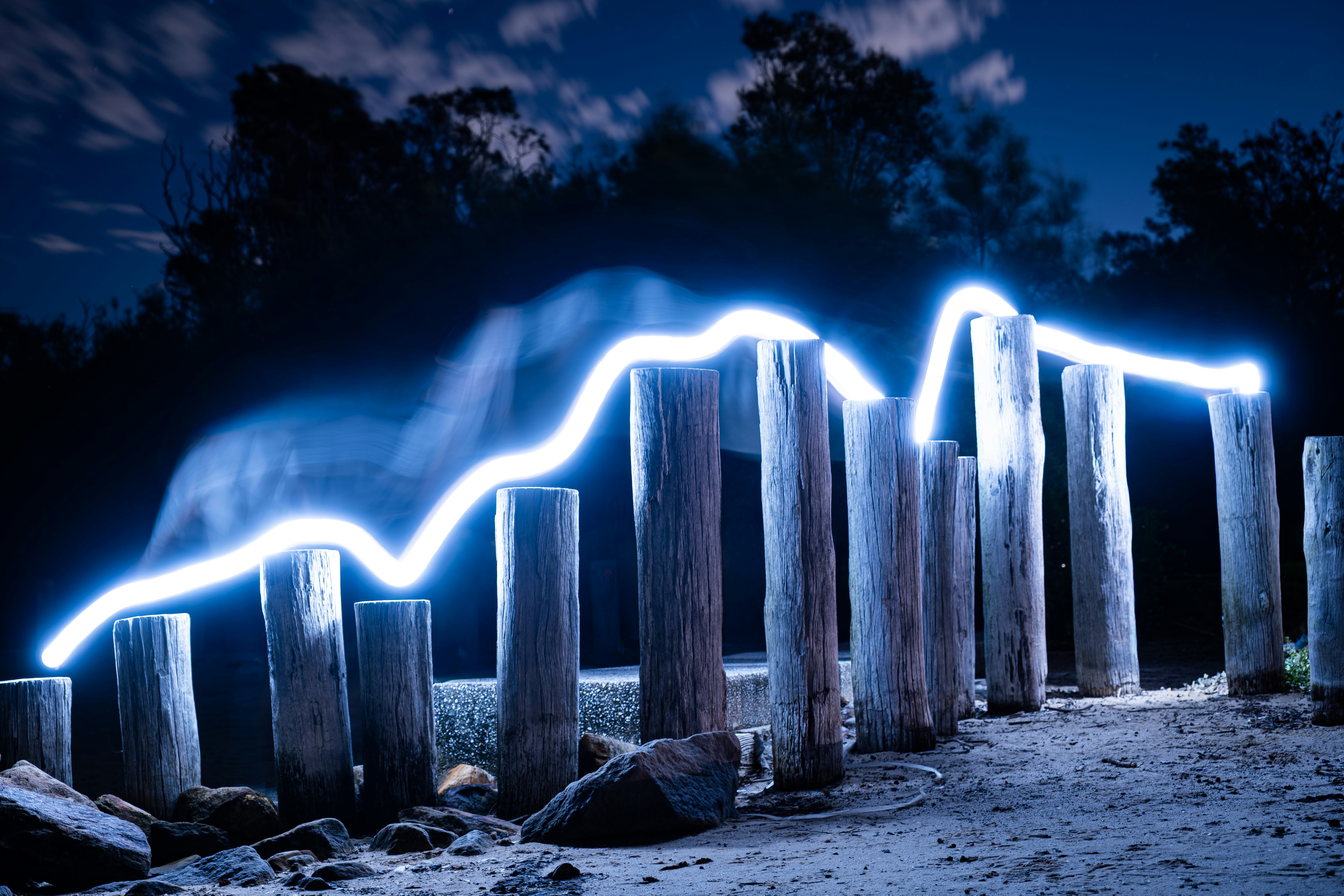 Long exposure night photo showing a bright light trail moving across a row of wooden posts on sandy ground with trees and clouds in the background.