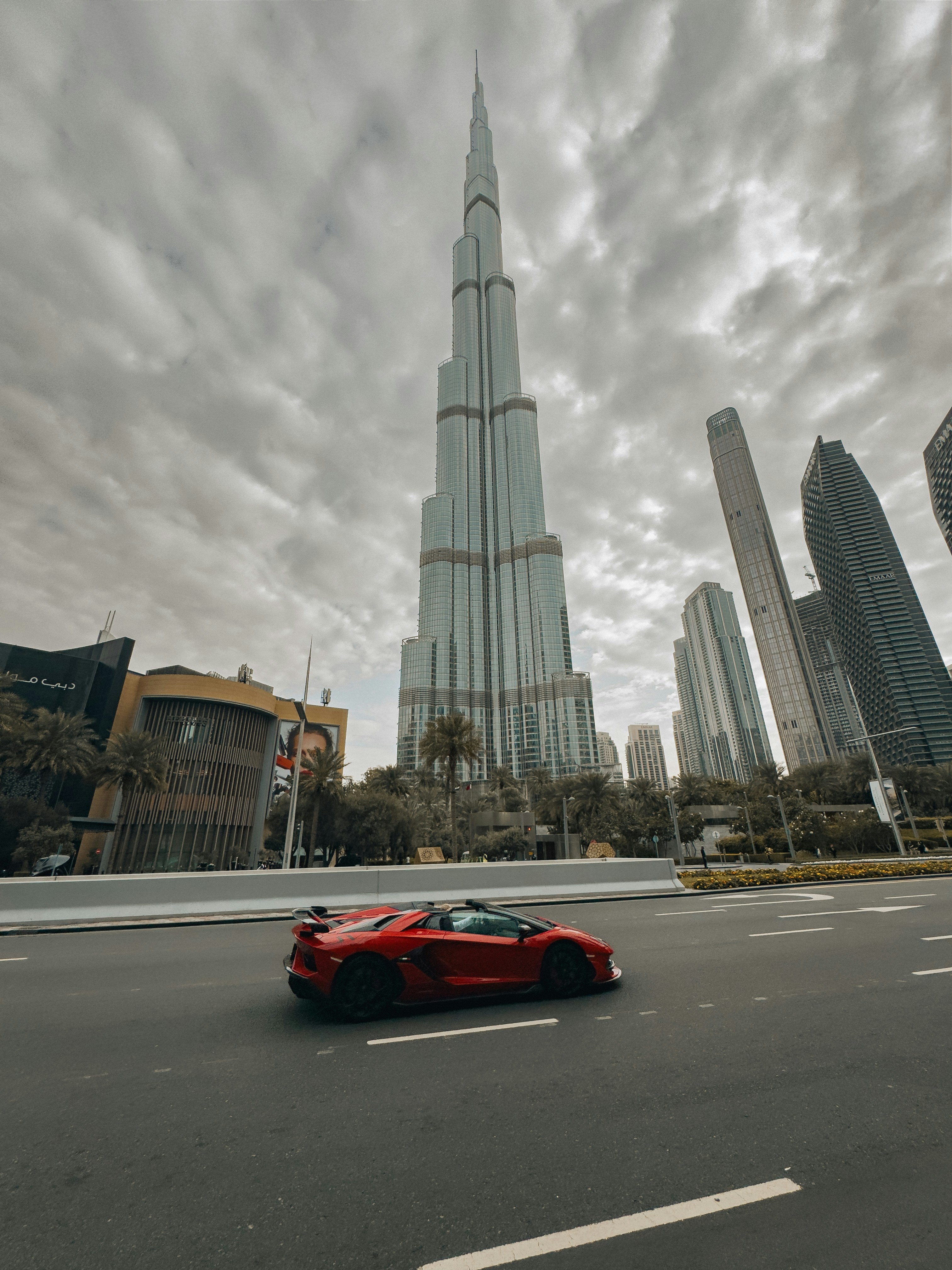 A dramatic street-level view of Burj Khalifa towering above Downtown Dubai, with a red sports car passing in the foreground under moody clouds.