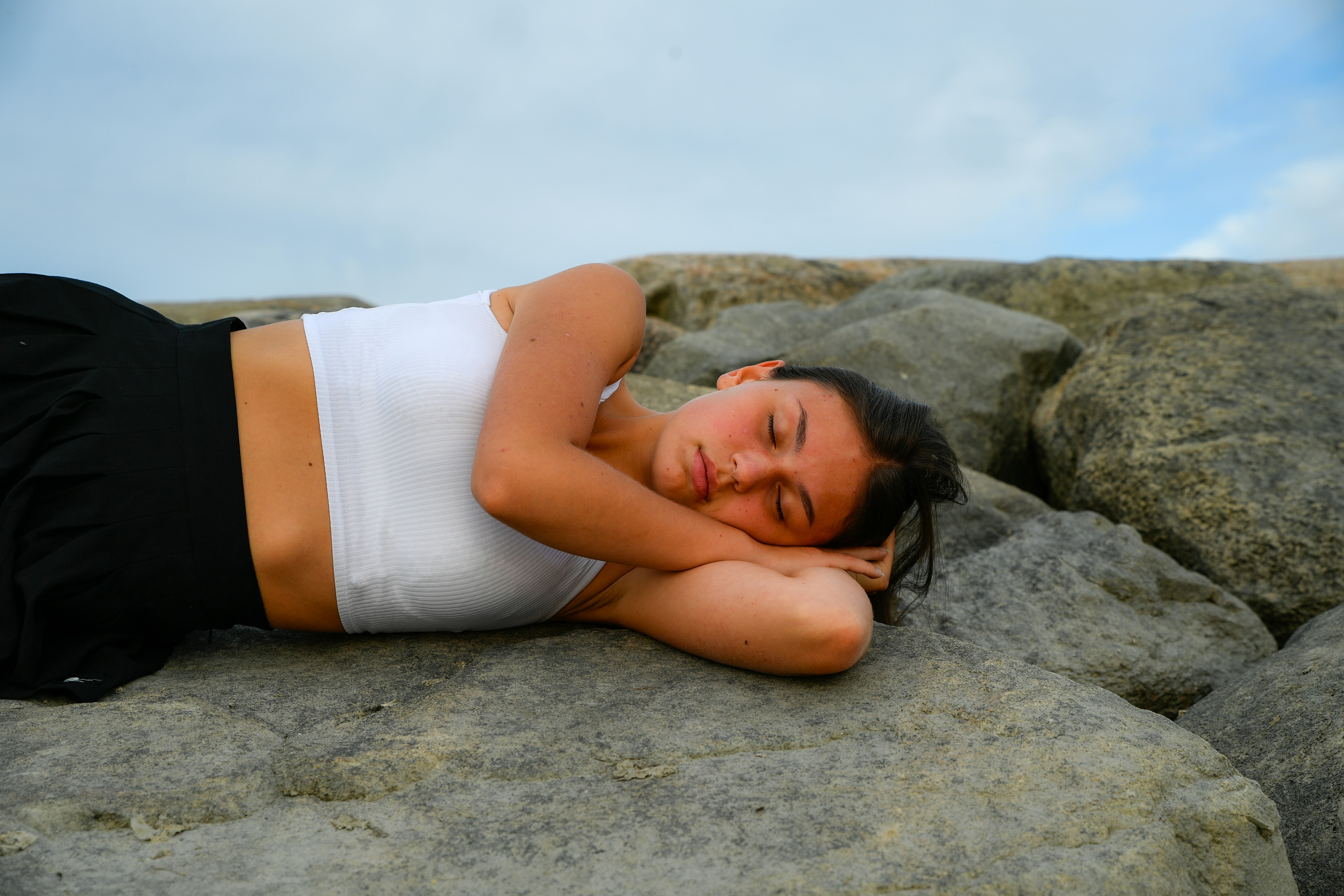 A young brunette woman in a black skirt and white top lying sideways on large seaside rocks in Pirita, Tallinn, with her eyes closed as if peacefully resting on a warm summer day.