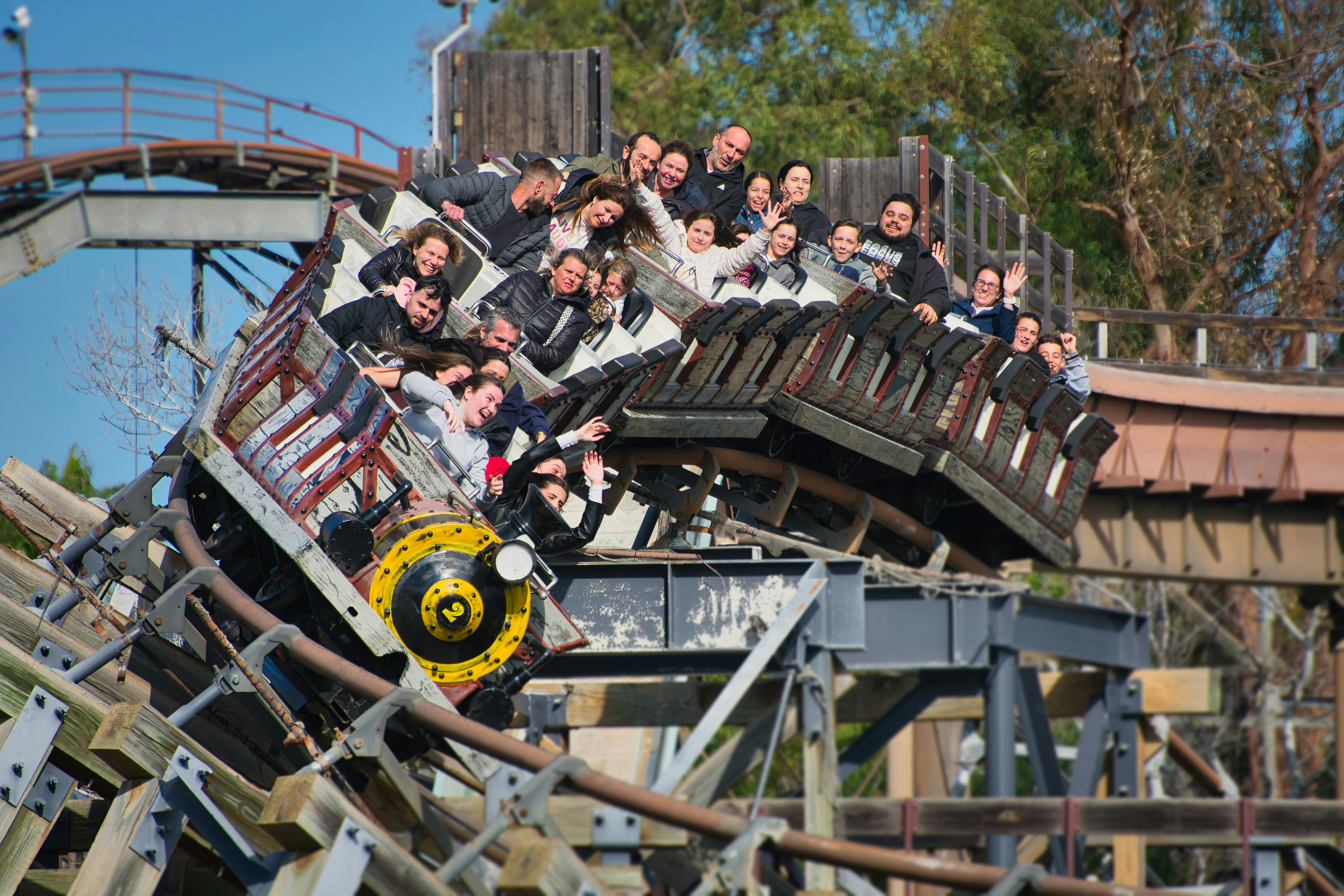 A crowded rollercoaster car tilts across a windy track with riders leaning outward in excitement; the image captures the mid-ride moment with a bright sky in the background. This is a photograph, not an illustration.