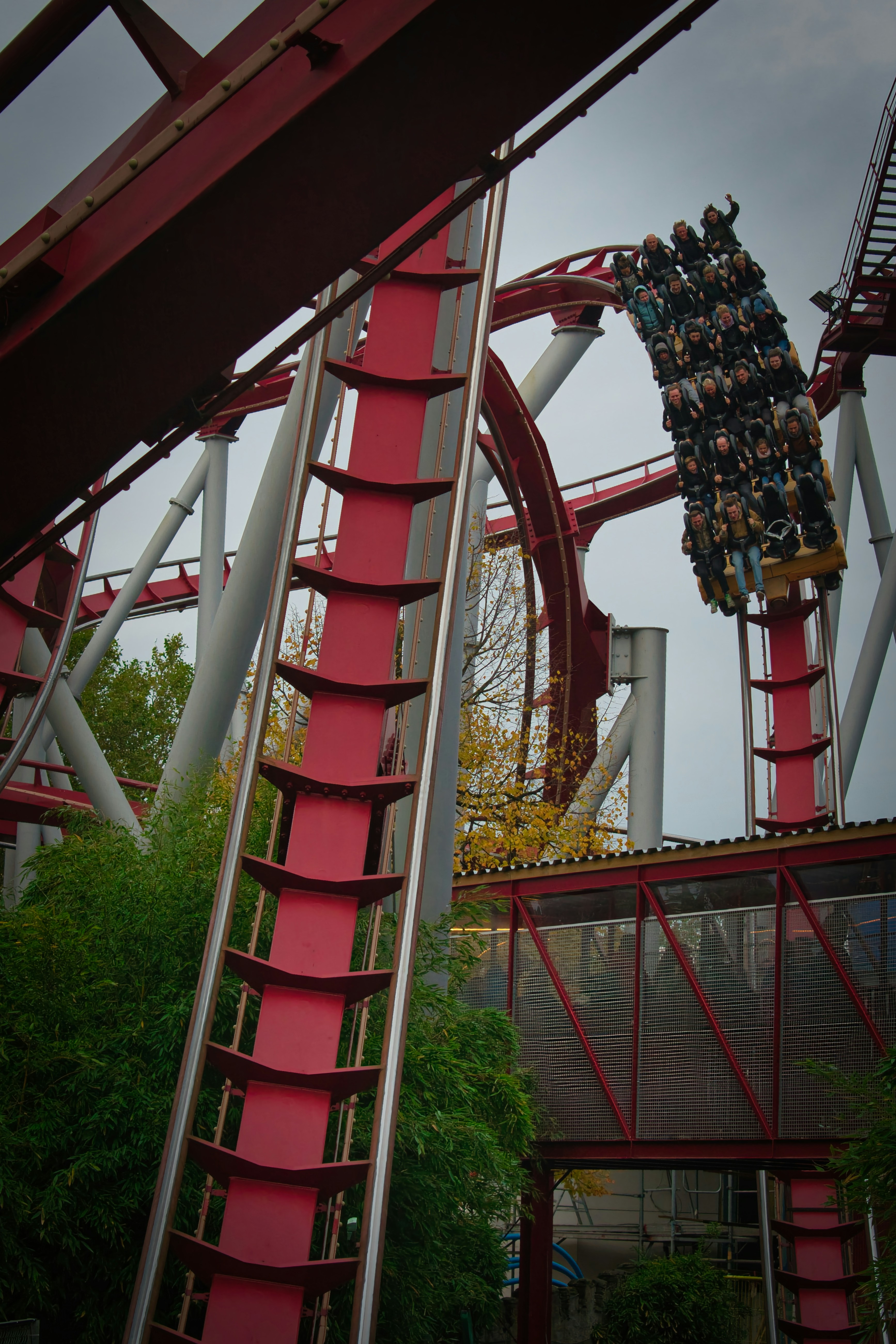 Roller coaster car twists through red rails with riders clinging to the loop on a gray sky day. A steel structure and green trees frame the scene, conveying motion and thrill.