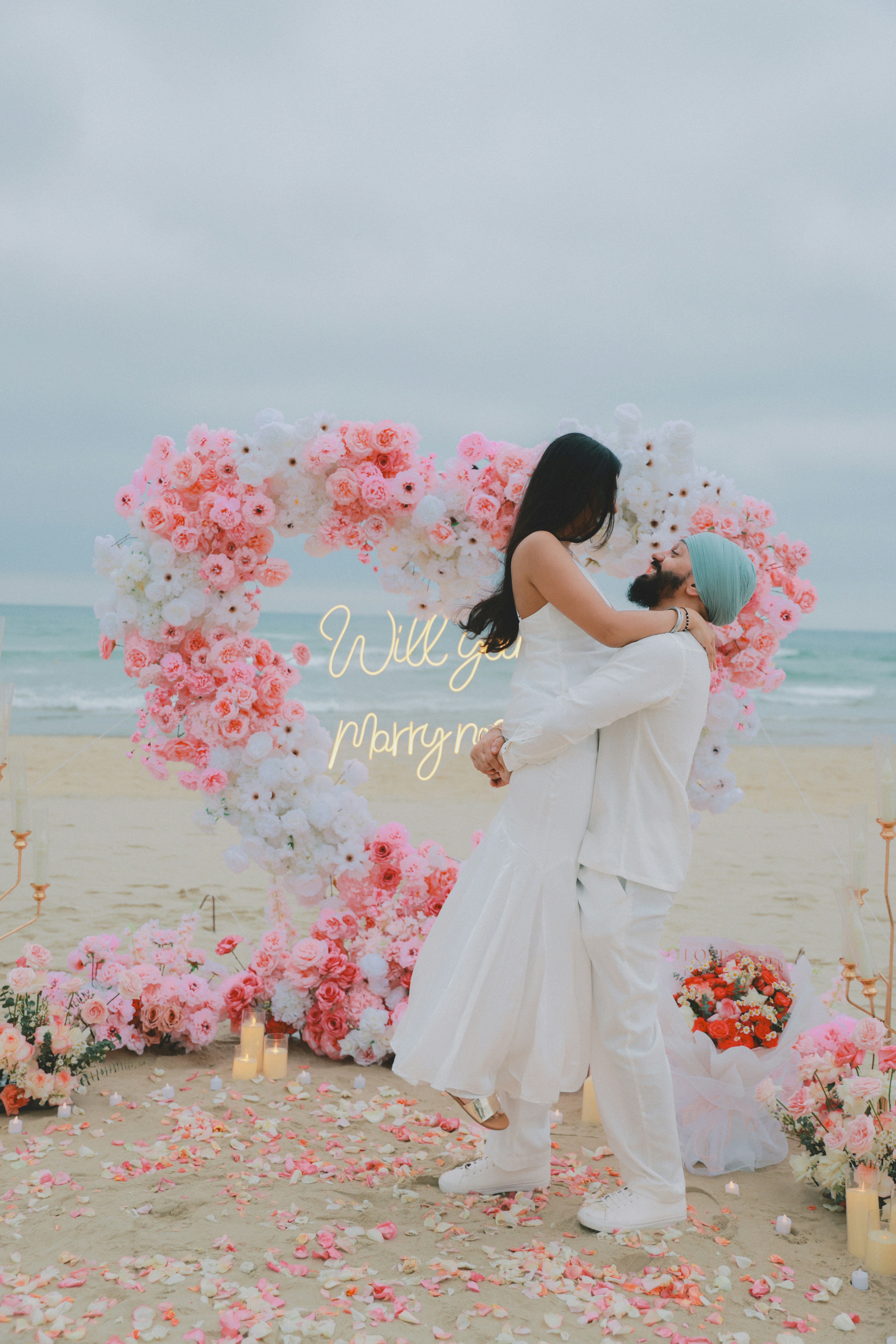 Beachside vow: a couple in white embrace before a heart of pink blooms