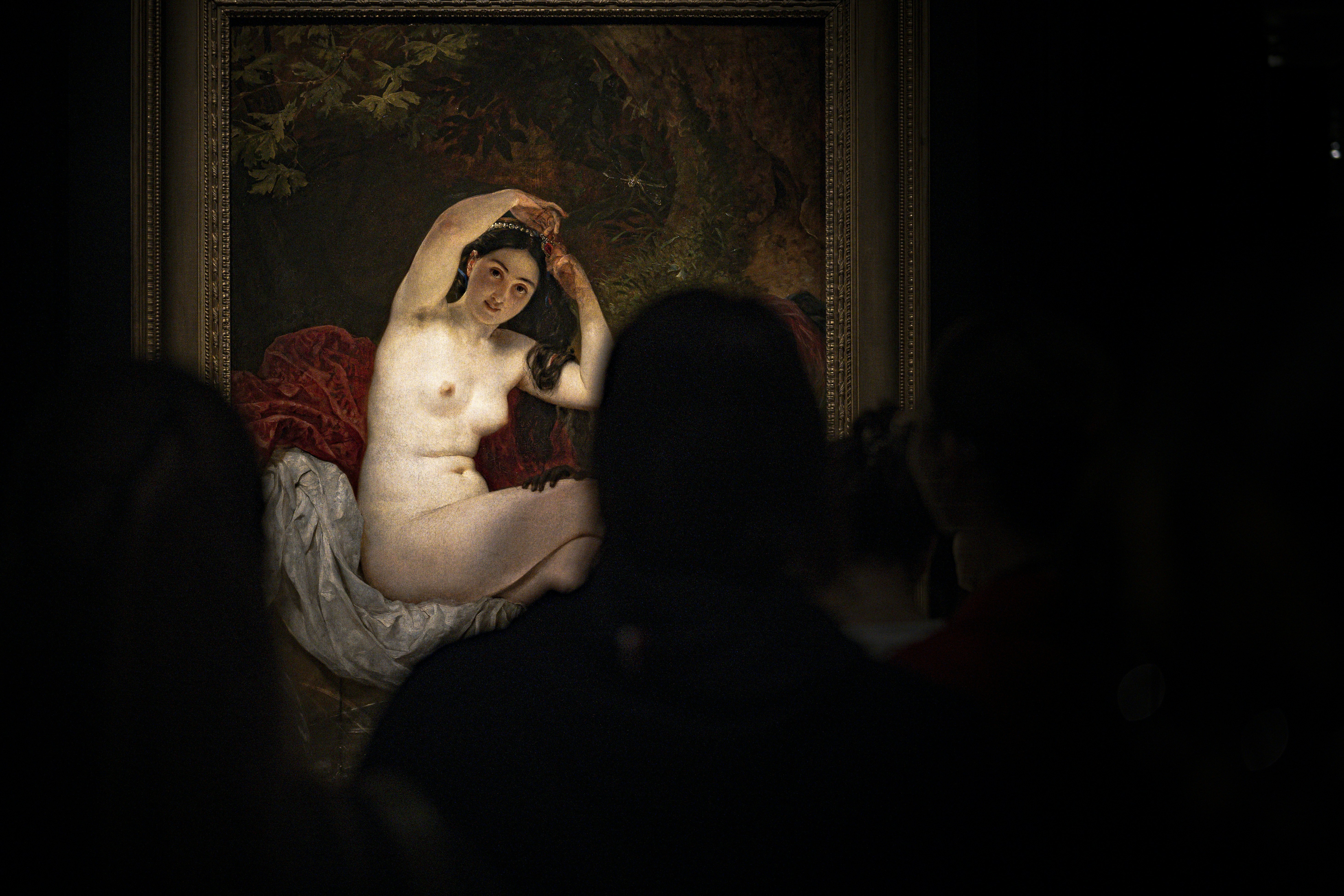 This evocative photograph captures a classic painting by Karl Bryullov exhibited in the New Tretyakov Gallery in Moscow. The glowing nude portrait is beautifully illuminated against a dark background, with the subtle silhouettes of viewers in the foreground. The scene radiates elegance, timeless beauty, and the intimate connection between art and its audience in a museum setting.