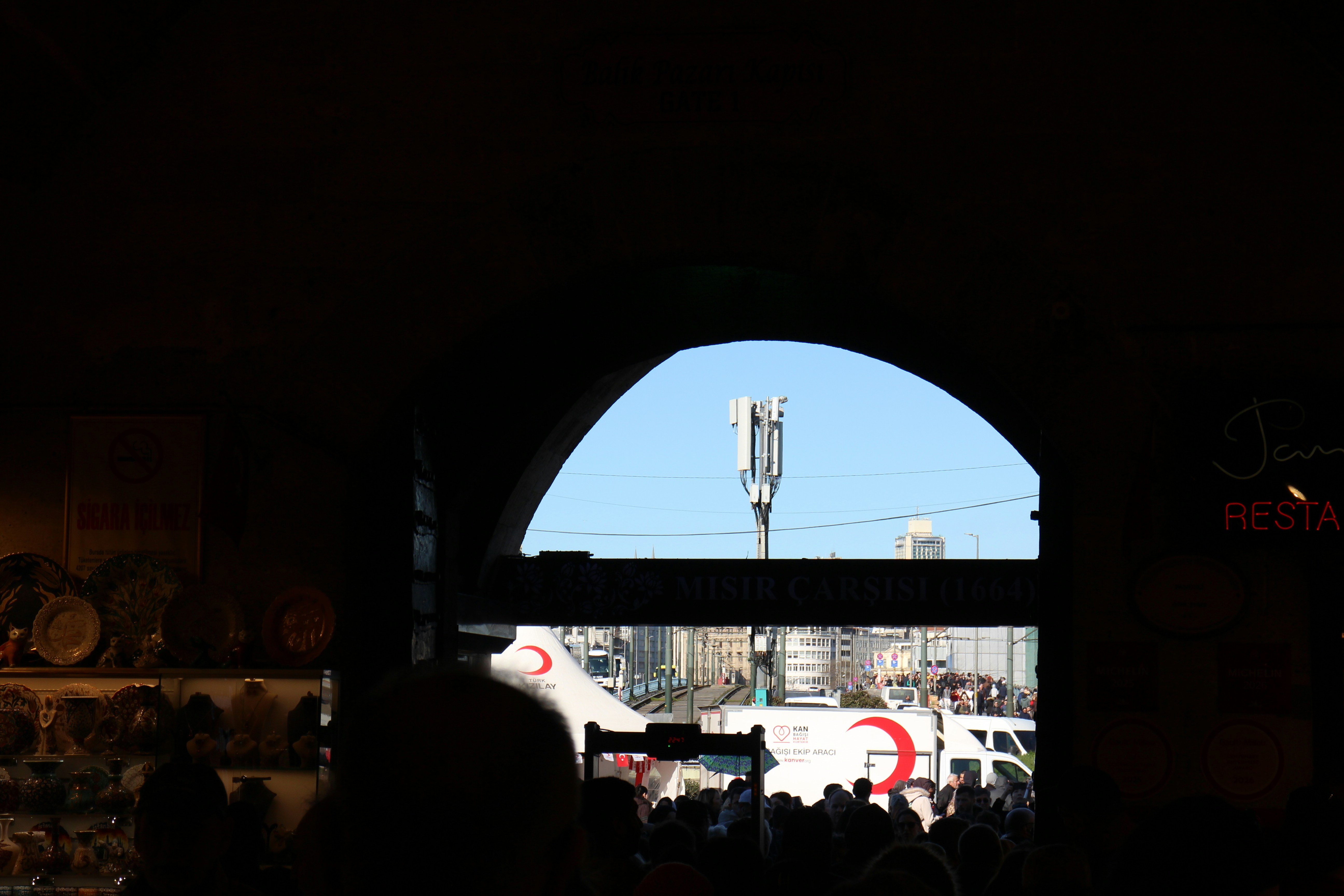 Dark interior framing opens to a bright city street beyond the arched doorway, with silhouettes of people inside and a distant skyline under a clear blue sky.