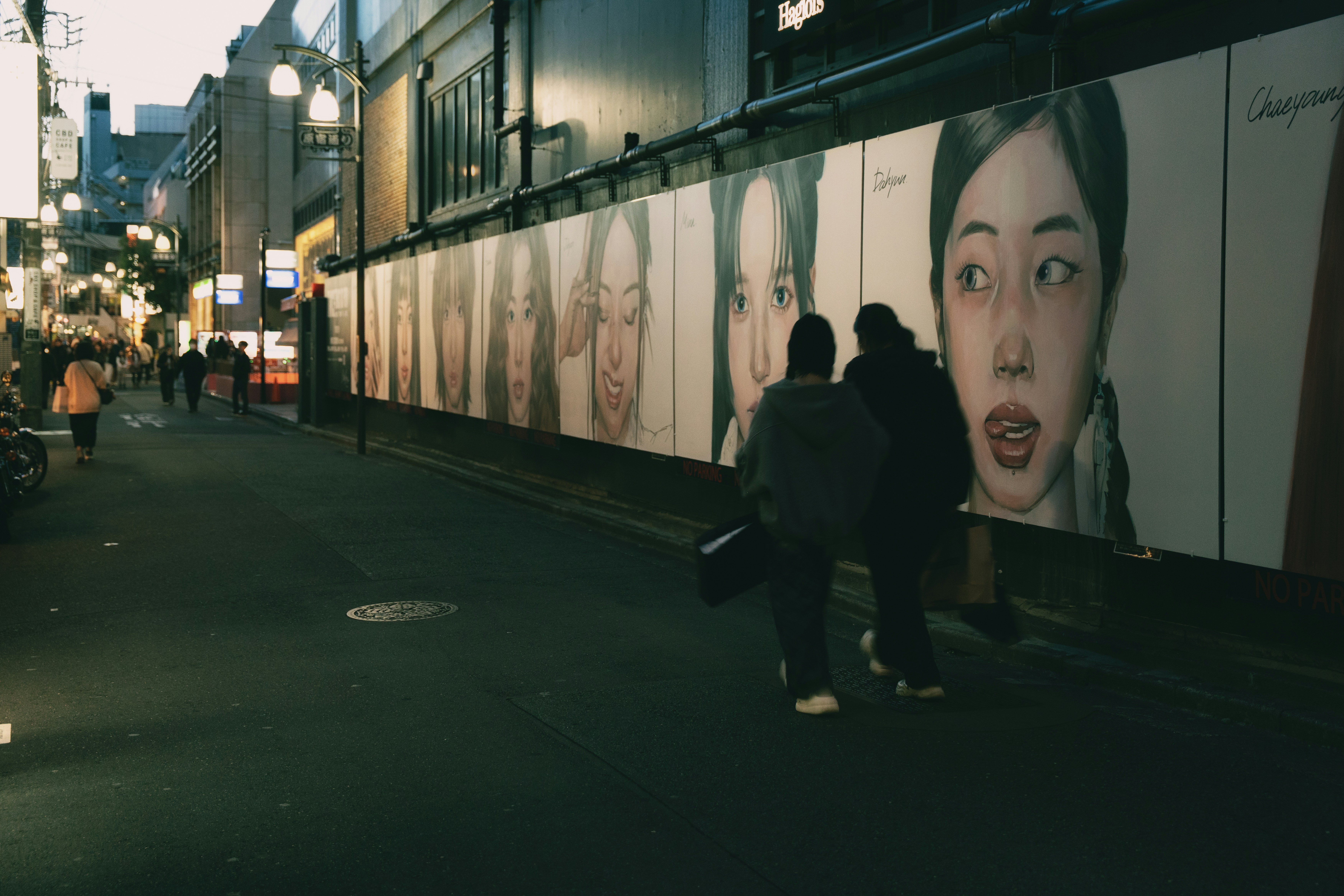 Two people walk past a mural of faces at night.