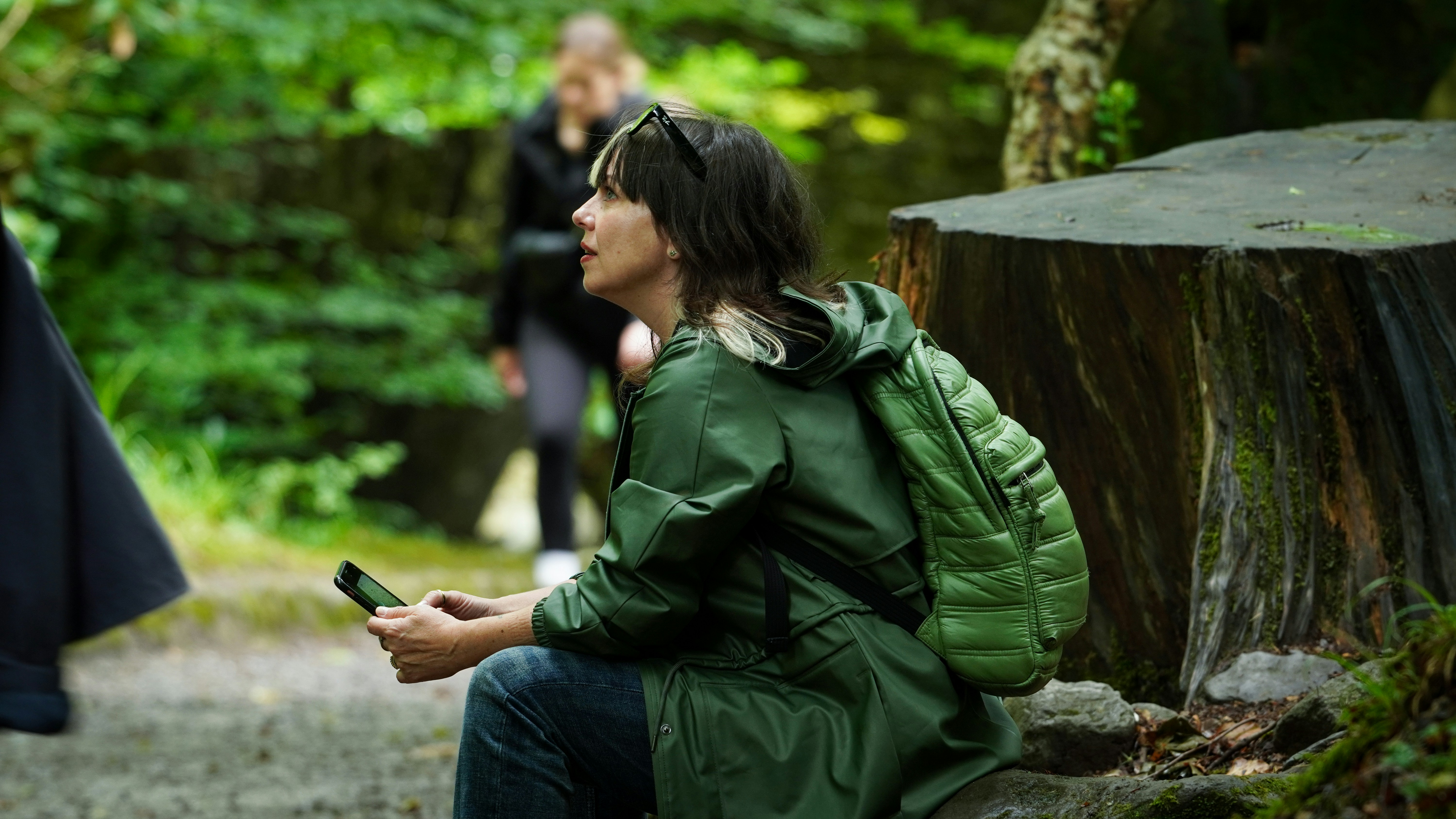 Green Jacket, Quiet Wait: A Woman with a Phone by a Forest Trail