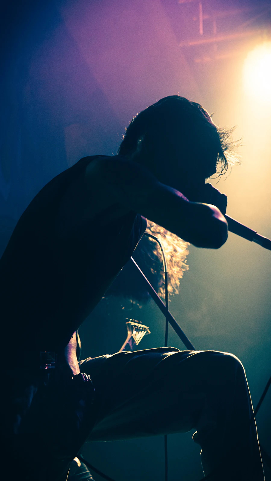 A dark-stage silhouette of a vocalist gripping a microphone, illuminated by contrasting warm and cool stage lights. The focus is on the performer's posture and presence, with the glow outlining the hair and microphone.