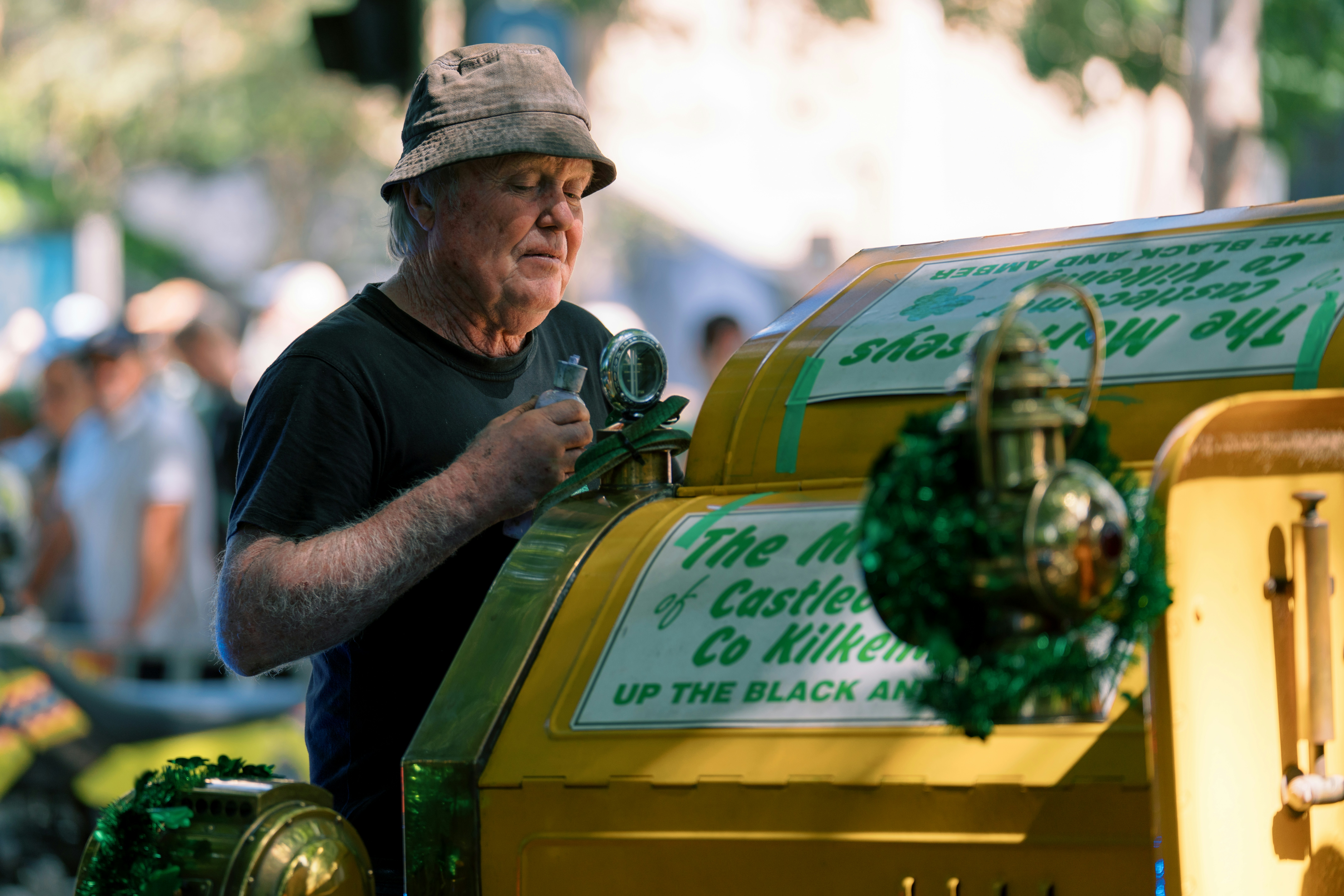 An elderly man in a hat examines a vintage yellow engine at a street parade, focusing intently with a small magnifier device in hand; the scene is framed by bystander blur and warm daylight. The setup suggests a public display or fair with a nostalgic mechanical exhibit.
