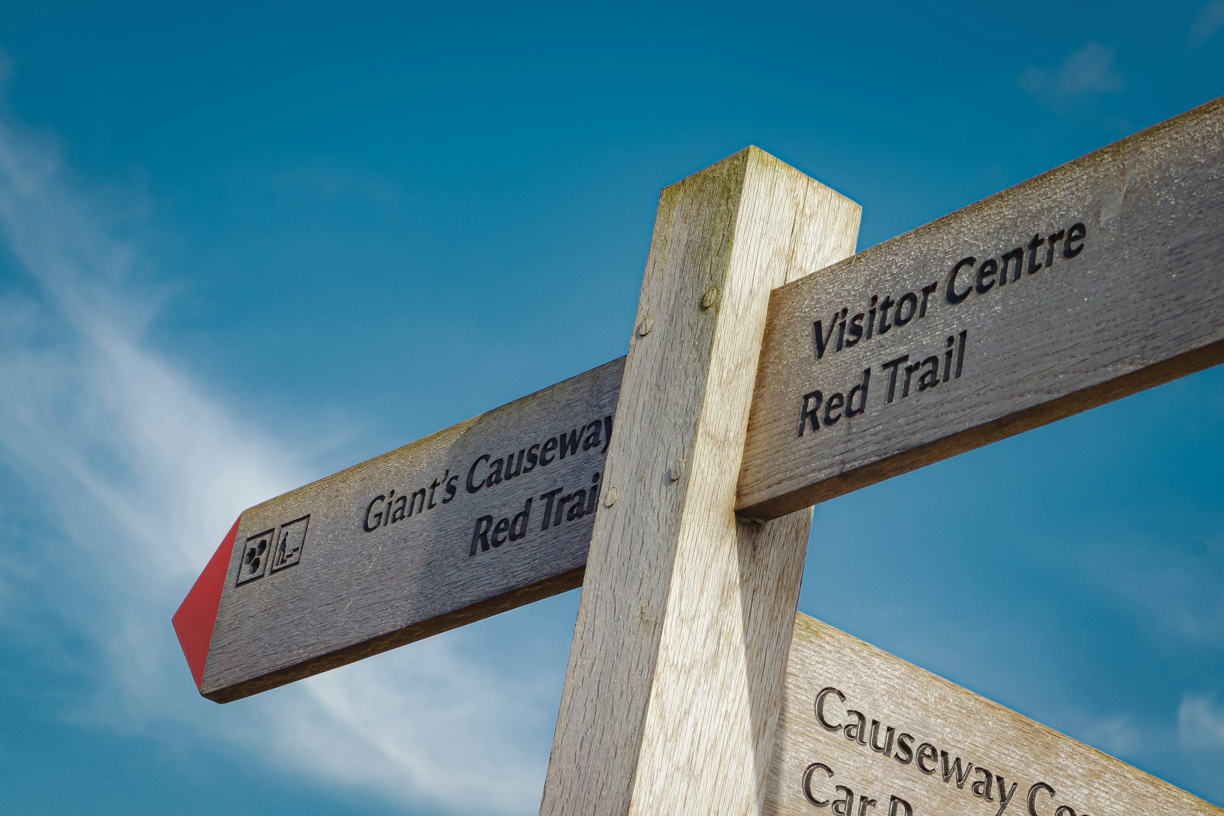 Wooden trail signpost with directional text against a bright blue sky, indicating routes such as Giant's Causeway Red Trail and Visitor Centre Red Trail.