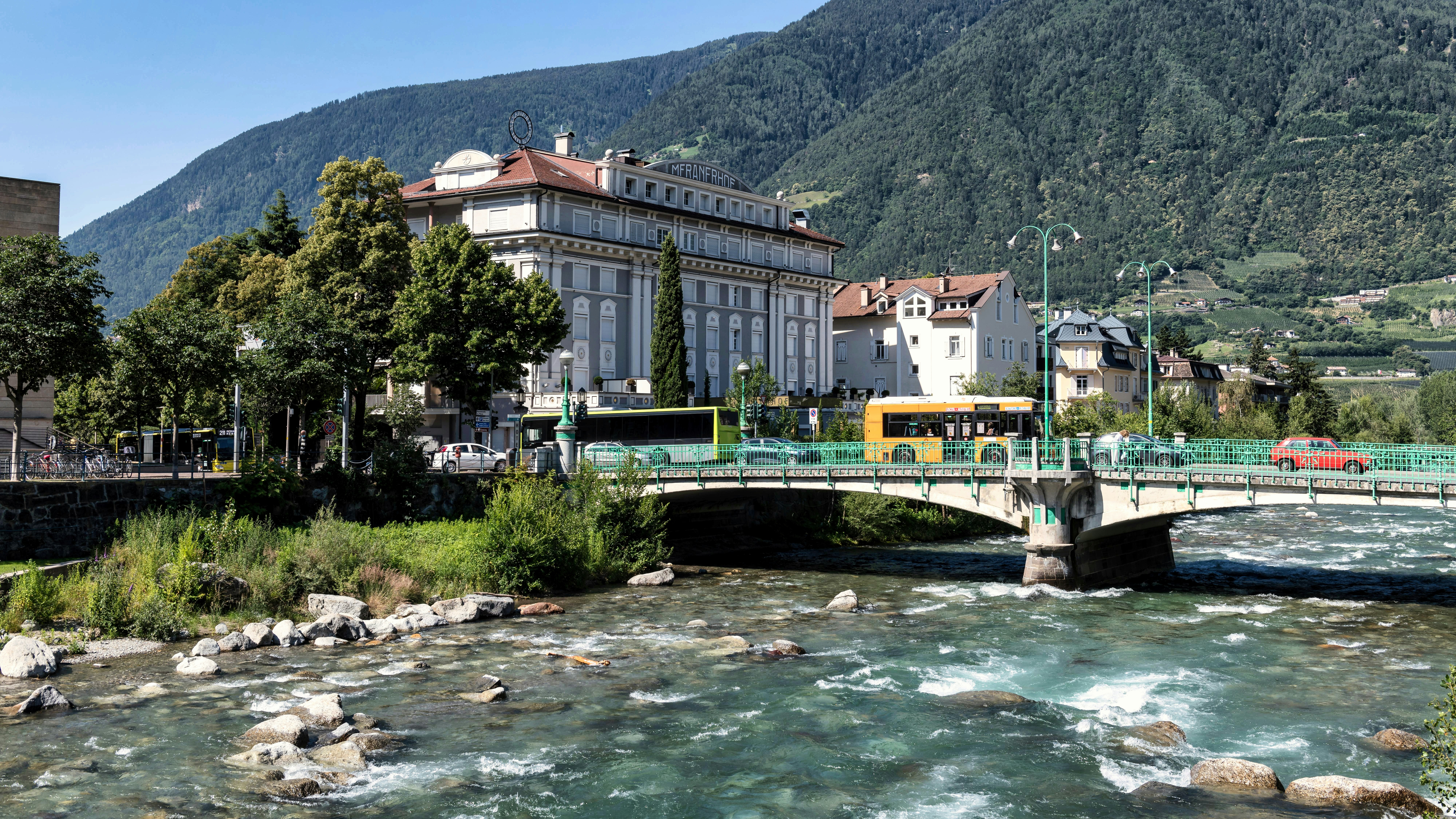 Stone-arched bridge over a lively riverscape with a grand town hall and surrounding hills