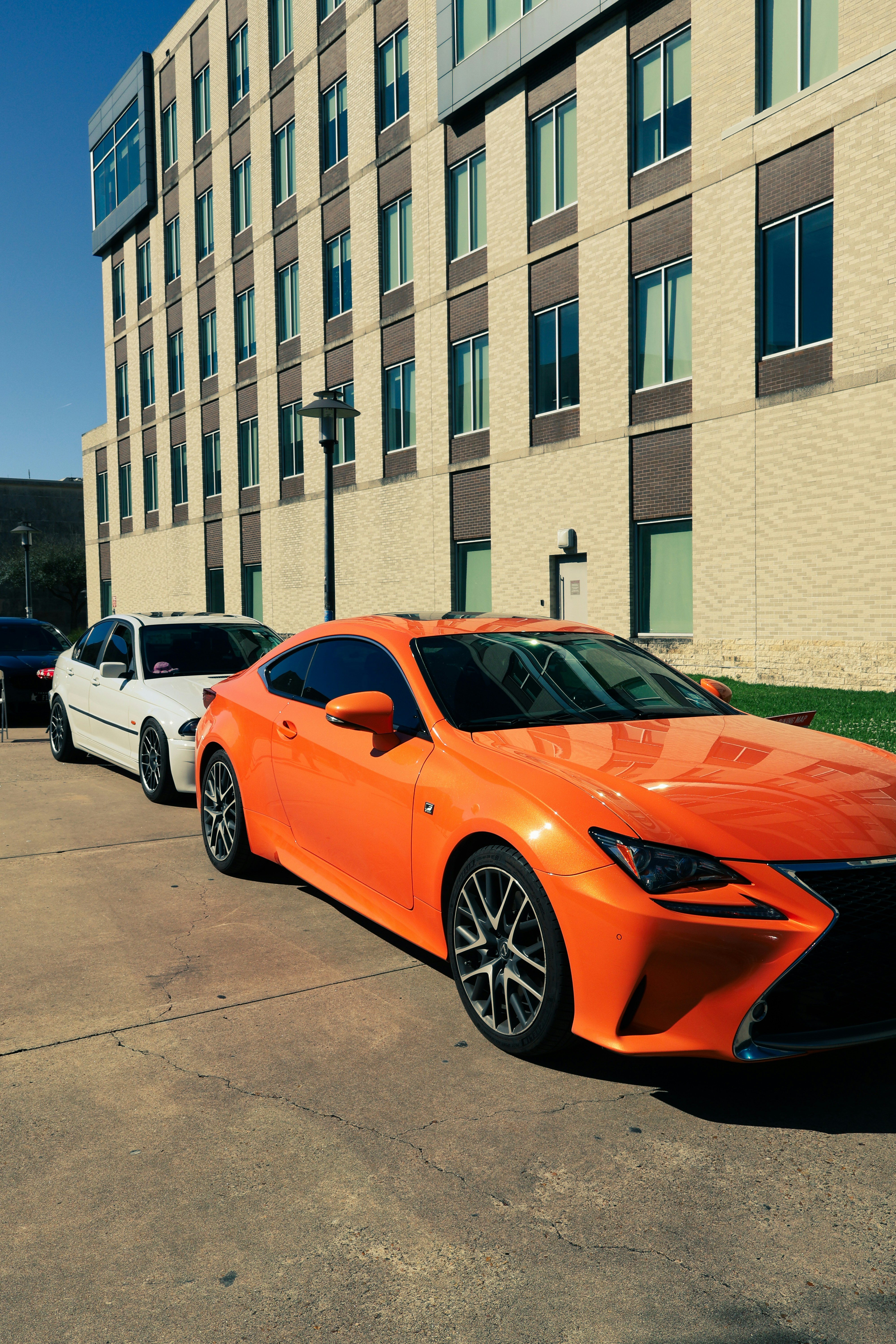 Orange sports car leads a sunlit line of sedans beside a brick campus building