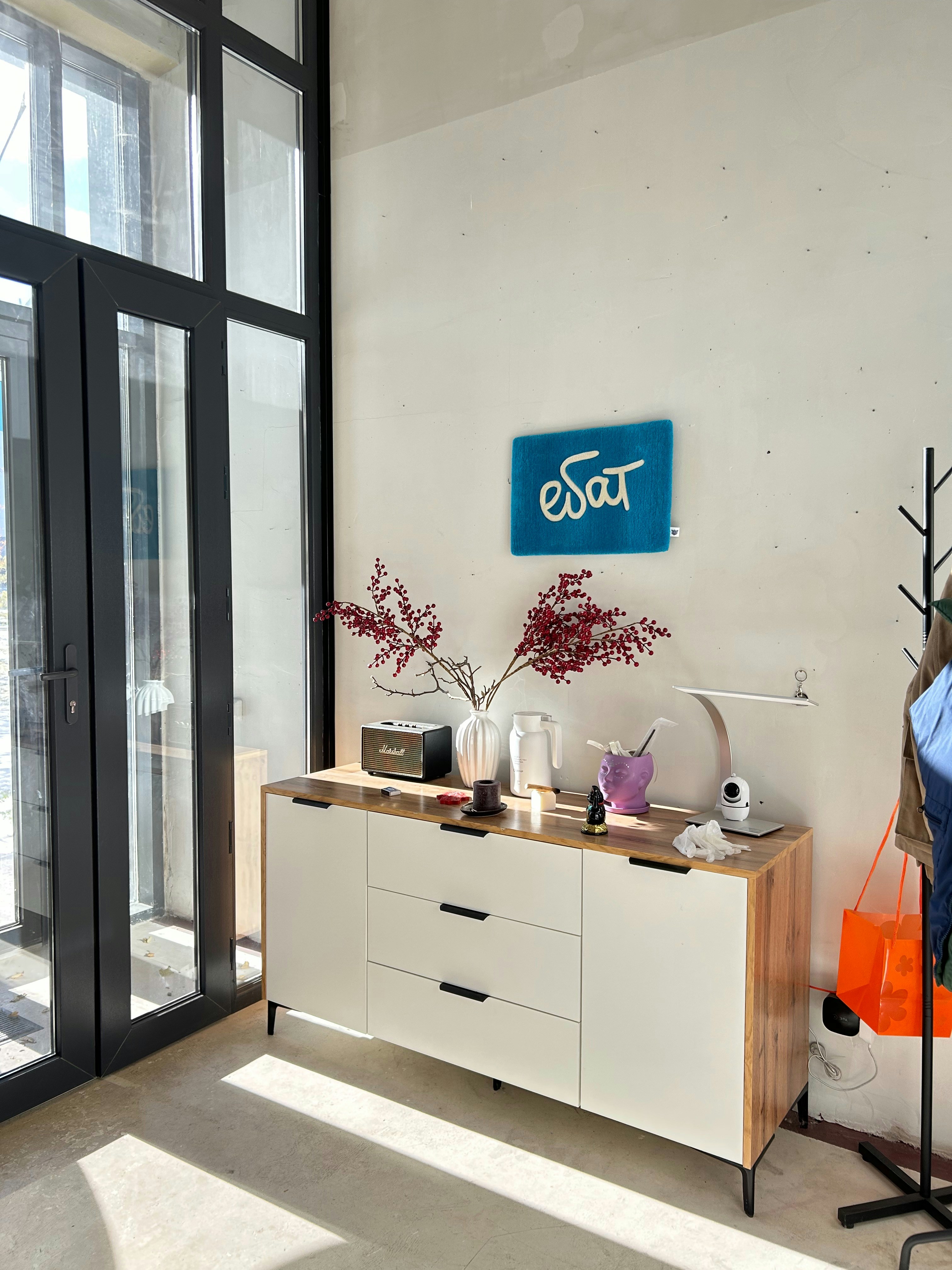 Bright interior with a two-tone sideboard under a blue abstract sign, red berry branches in vases, and a floor-to-ceiling glass entrance; a clean, modern setup focusing on natural light and minimal aesthetics.