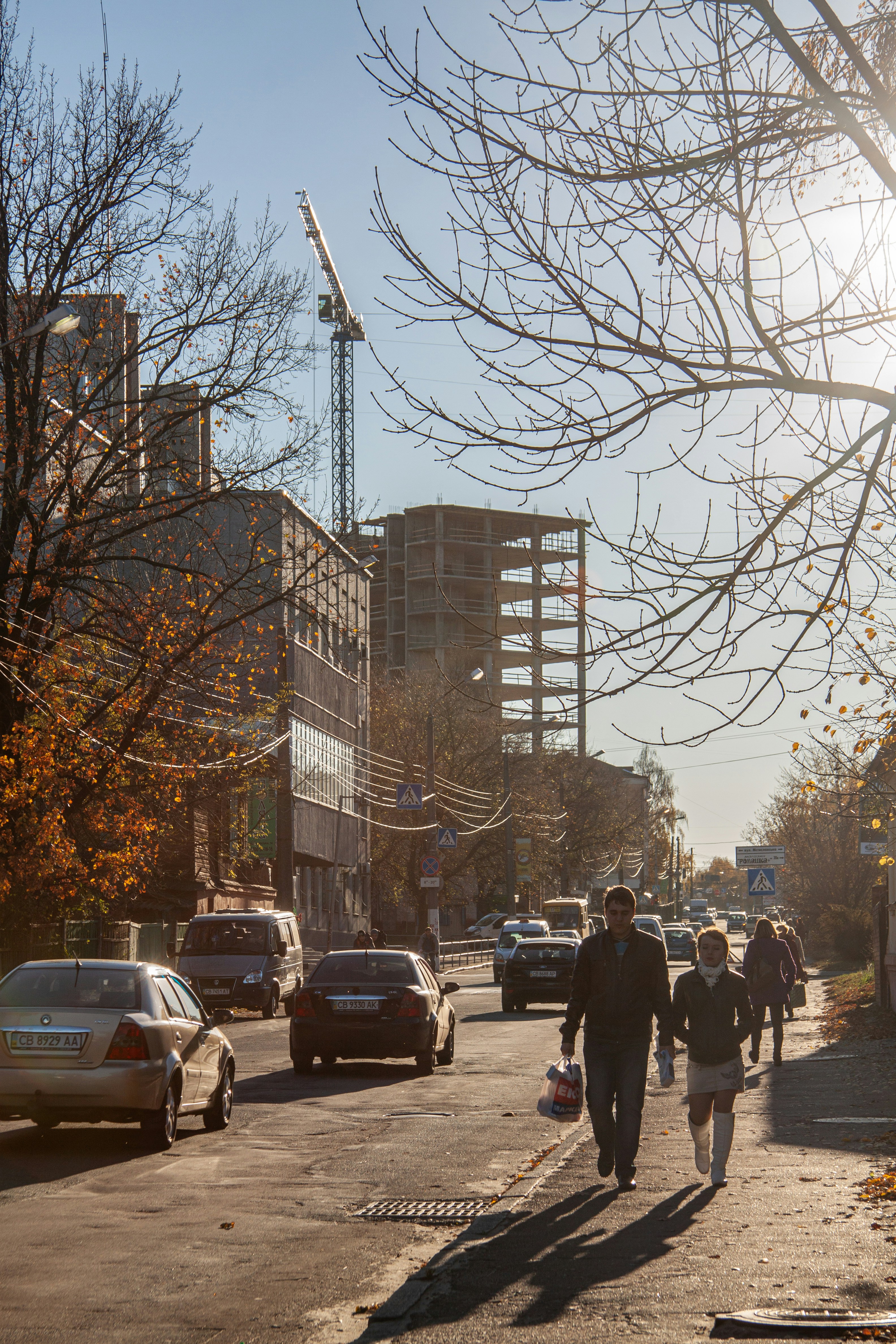Two pedestrians stride along a sunlit autumn street beneath a skeletal crane and leafless trees