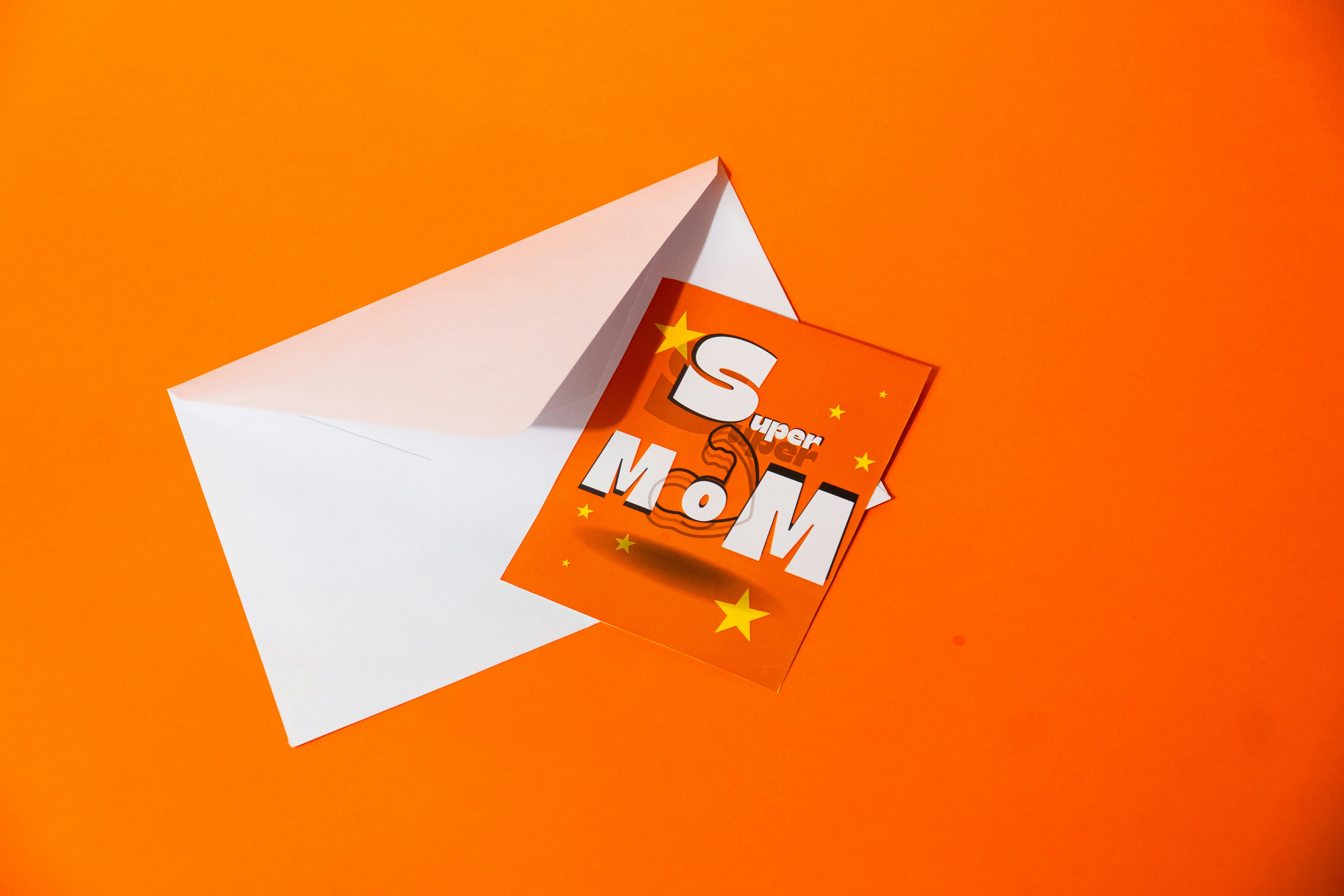 Orange backdrop, white envelope, and a bright card reading 'SUPER MOM' drifting from the flap