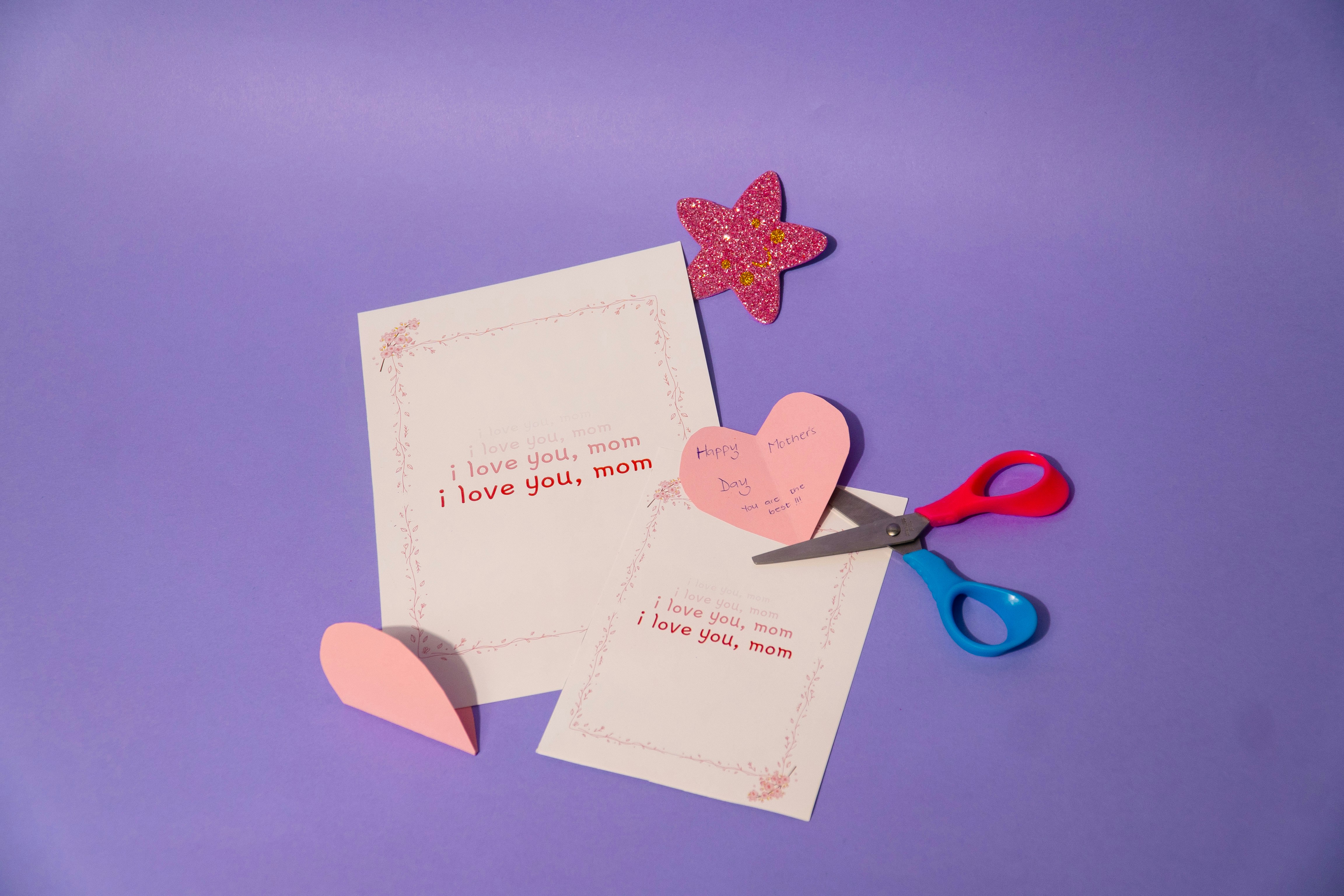 Two pink cards with printed messages lie on a purple background, accompanied by a red and blue pair of scissors and decorative paper hearts; a small glittery flower sits nearby, suggesting a craft moment.