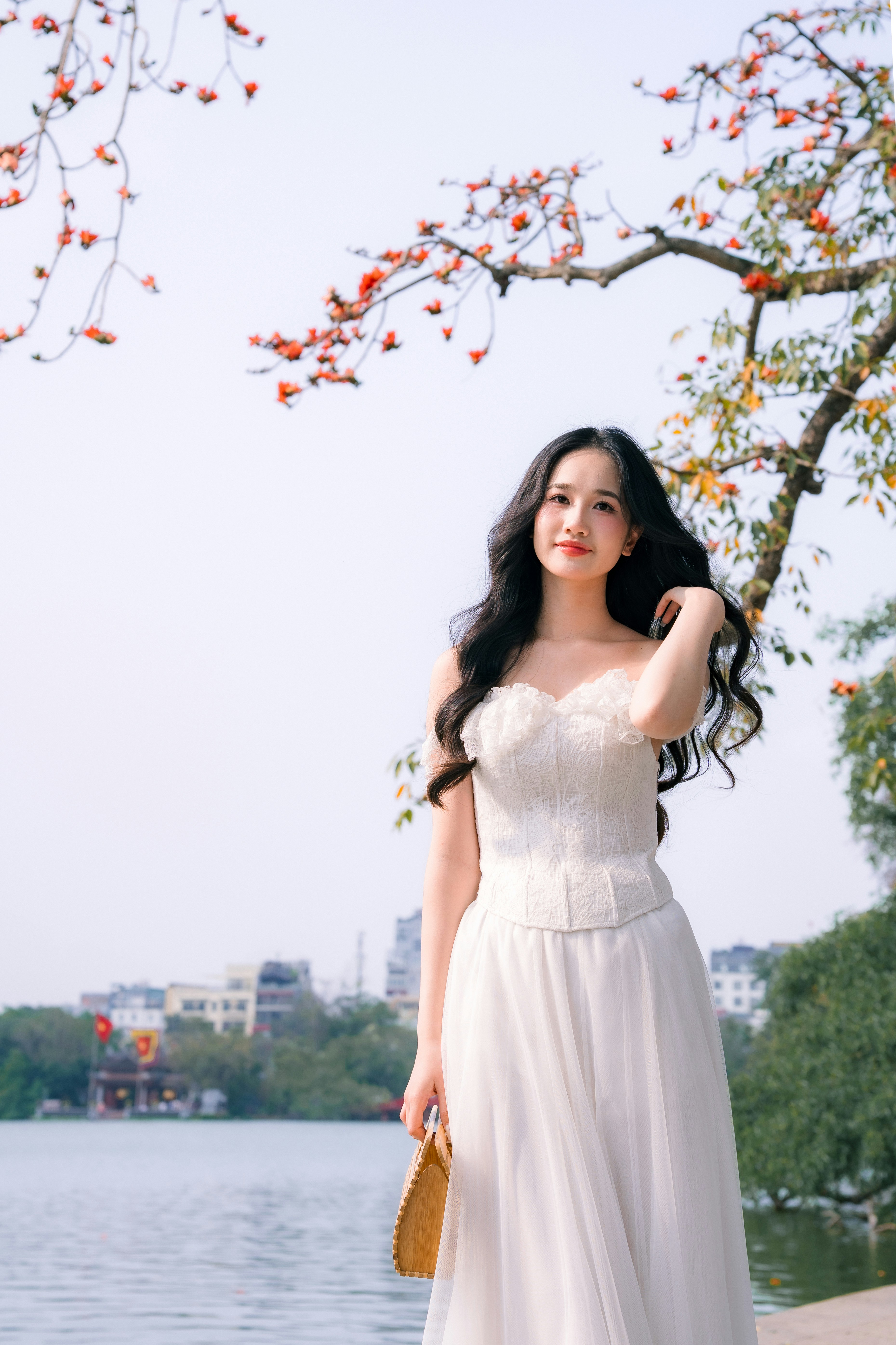 A serene portrait of a young woman in a white dress by the lakeside, surrounded by blooming flowers in Hanoi.