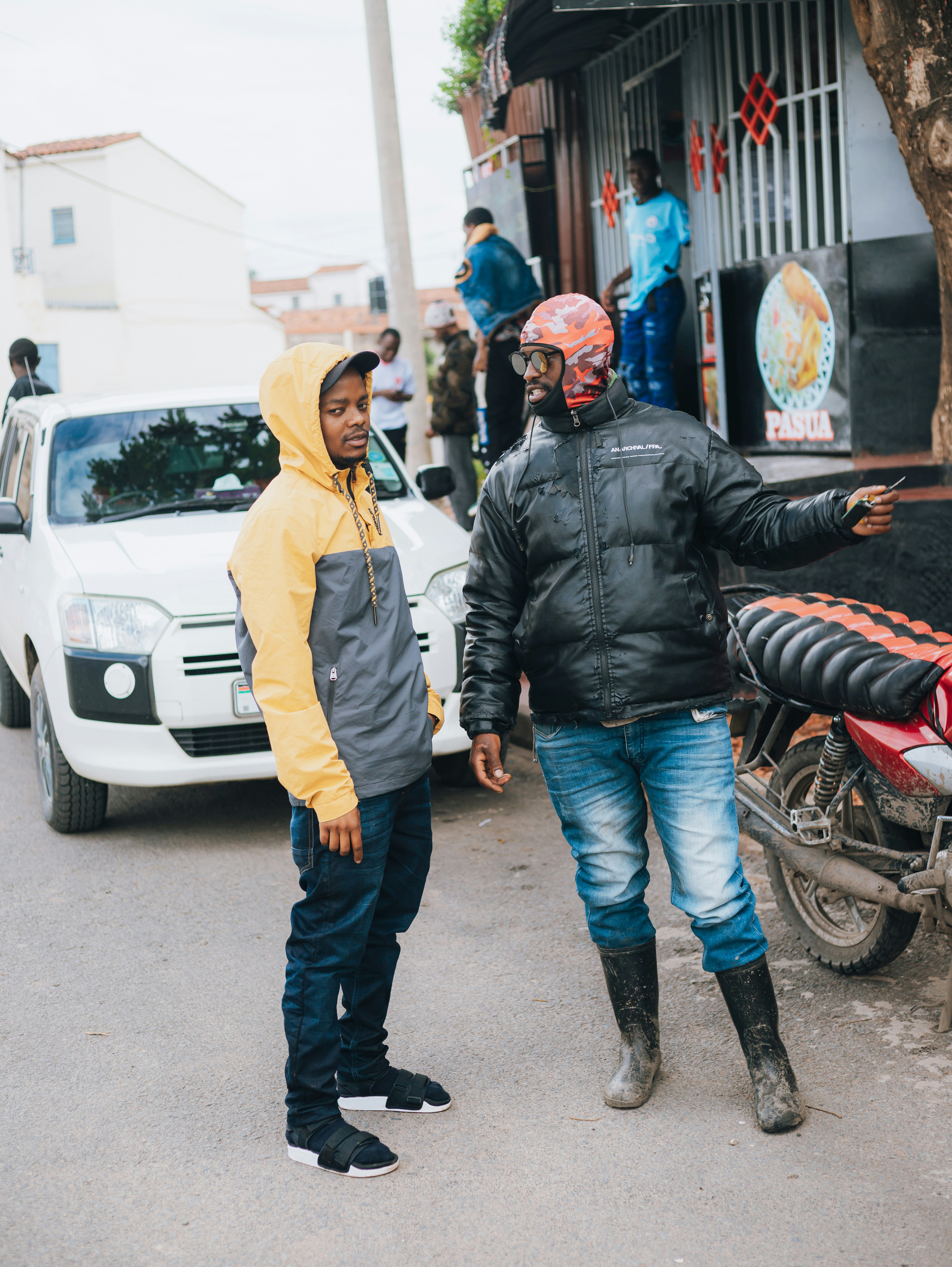 Two riders paused on a sunlit street, jackets and helmets catching the eye amid a casual urban scene