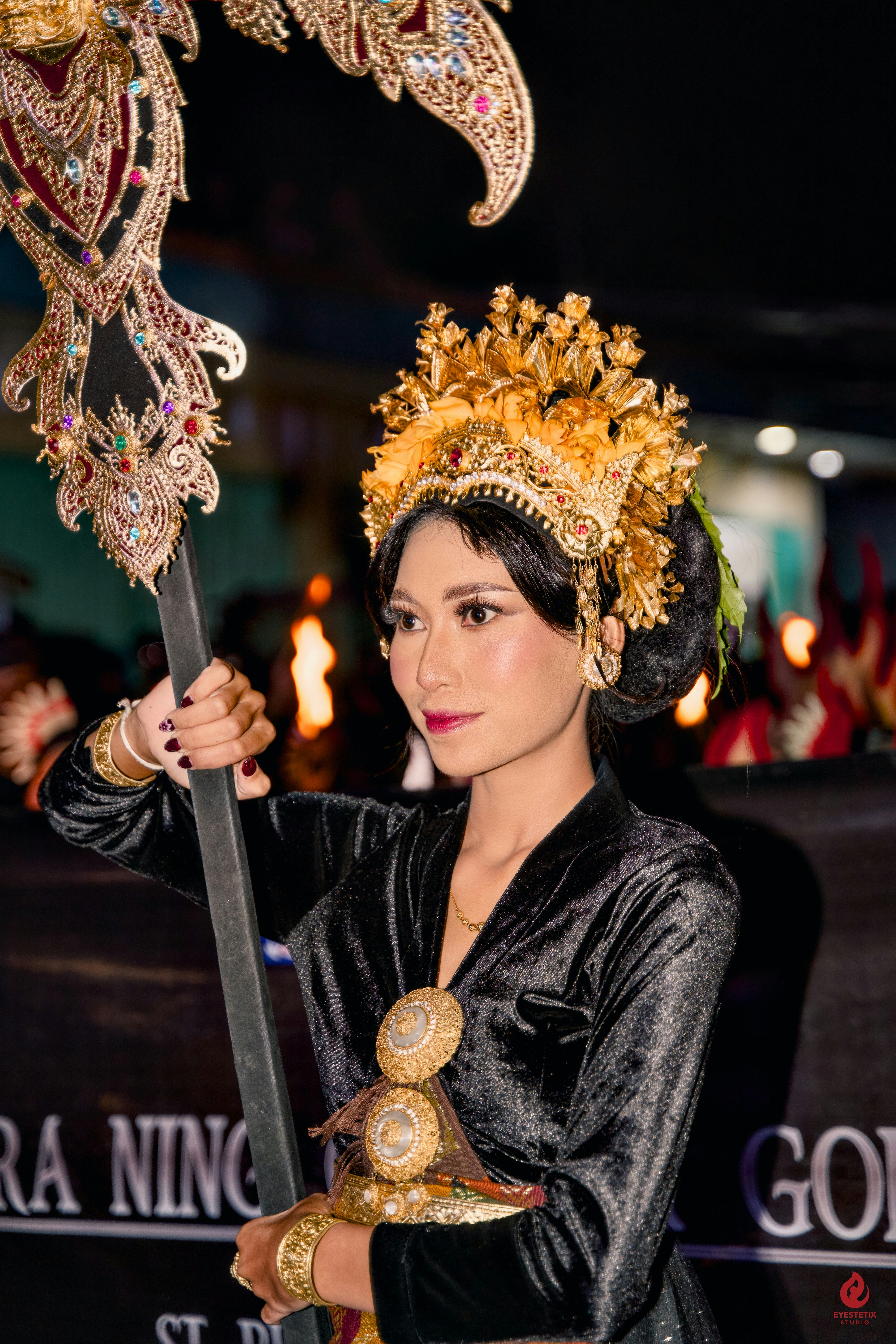 Regal performer in ornate headpiece wields ceremonial staff under warm night glow