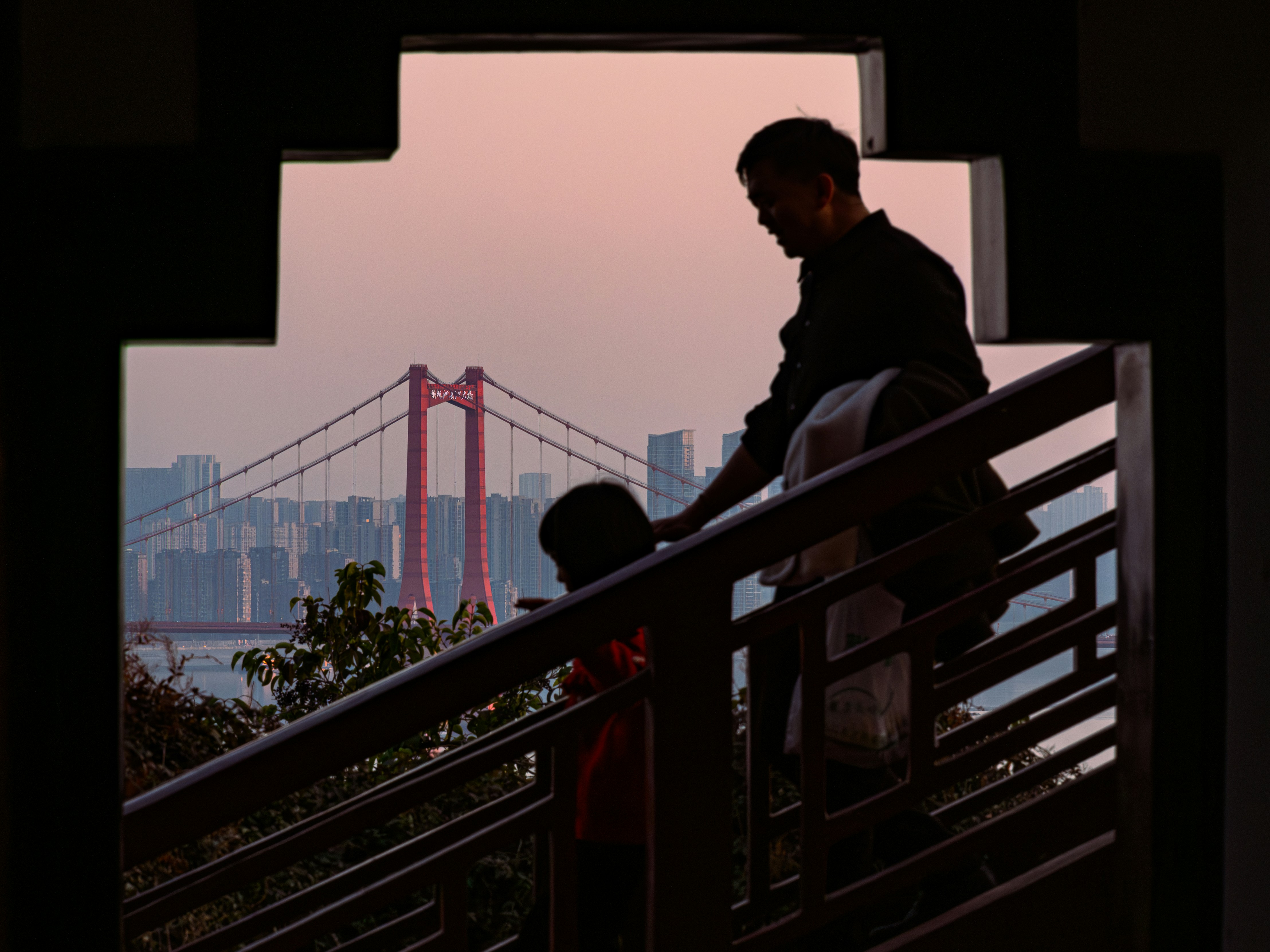 Silhouetted Figures on a Staircase Framing a Red Suspension Bridge at Dusk