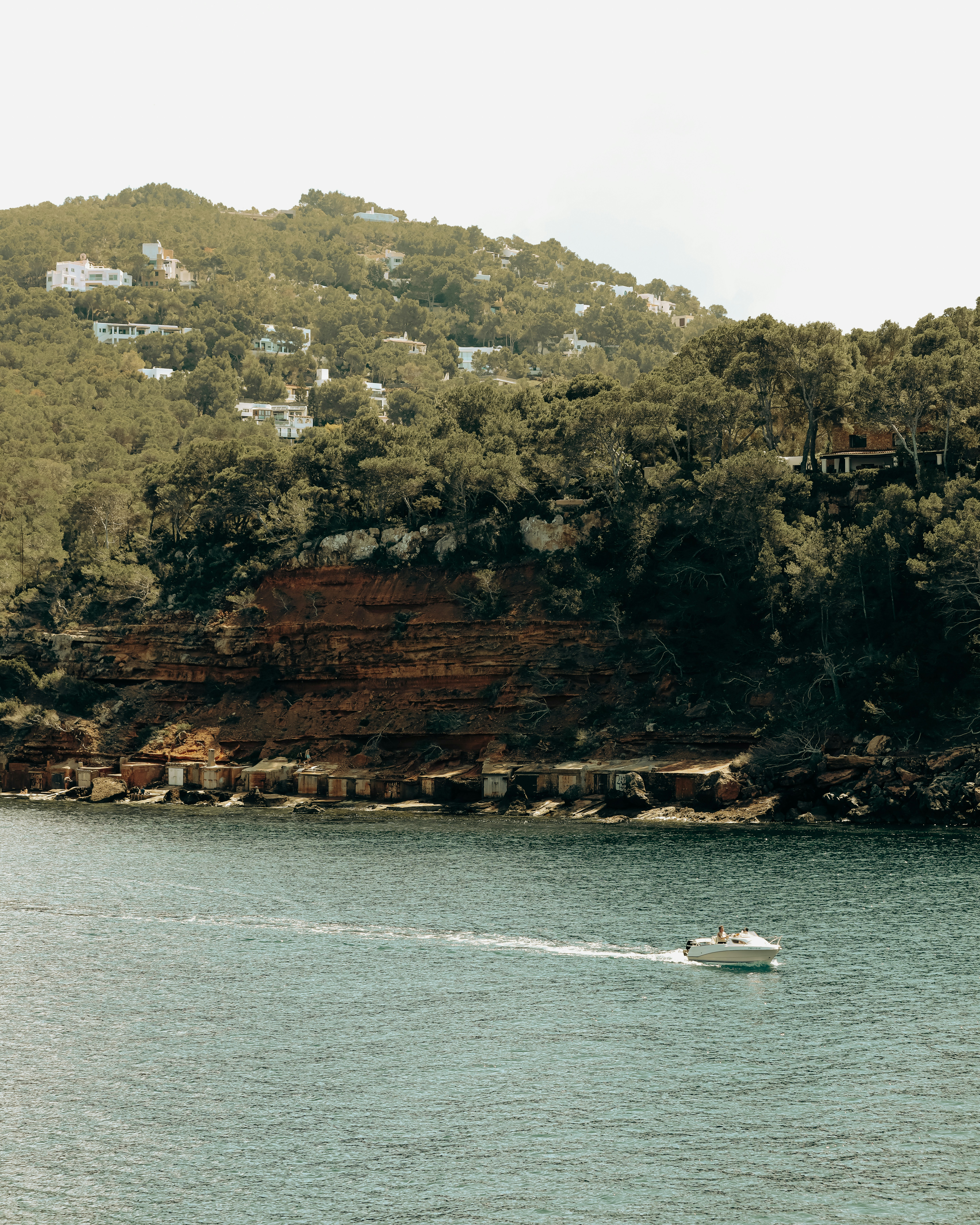Small motorboat glides along calm water near a tree-covered cliff with houses perched above.