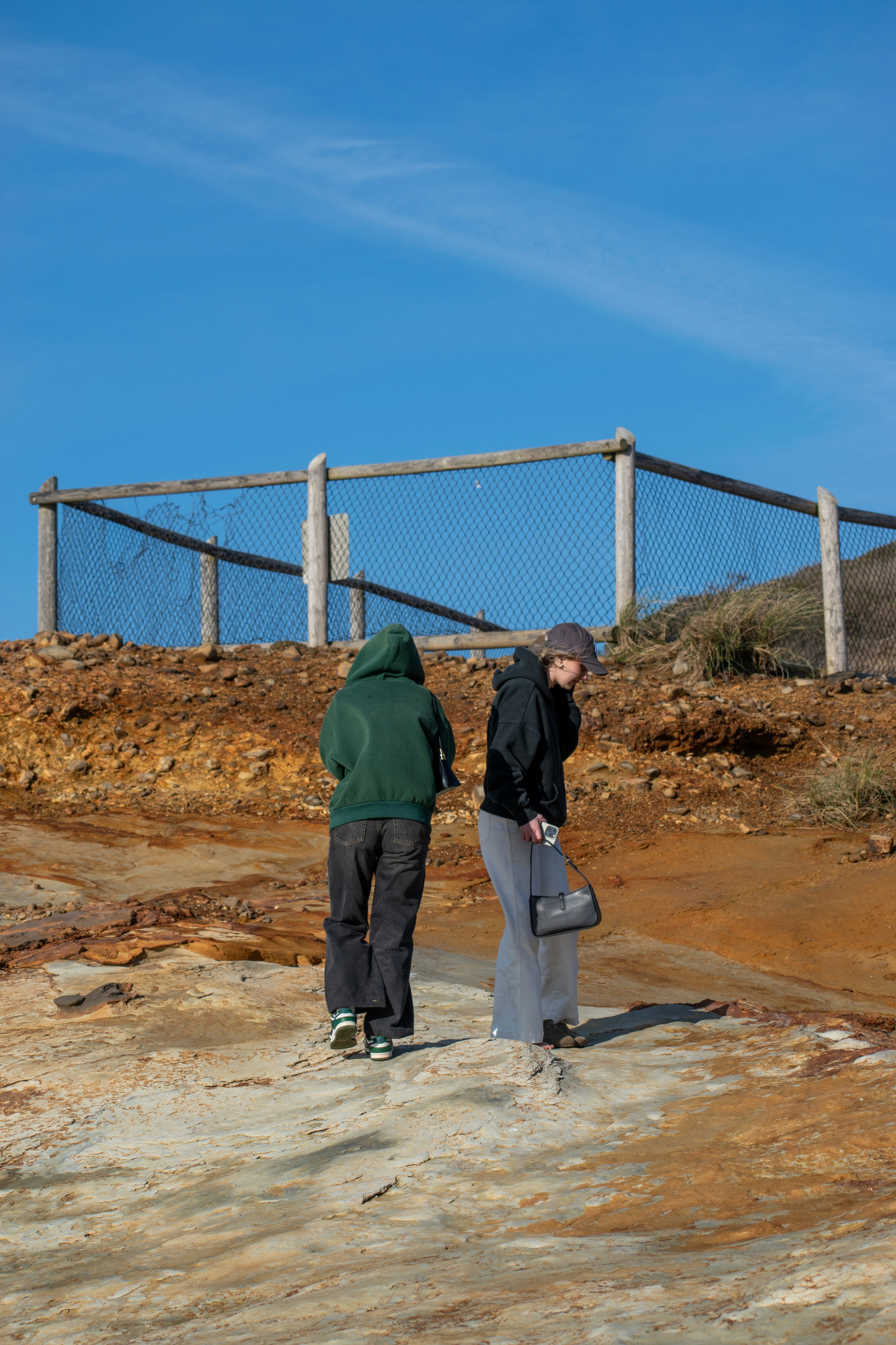 Two hikers pause near a weathered fence atop mineral-streaked ground under a clear blue sky