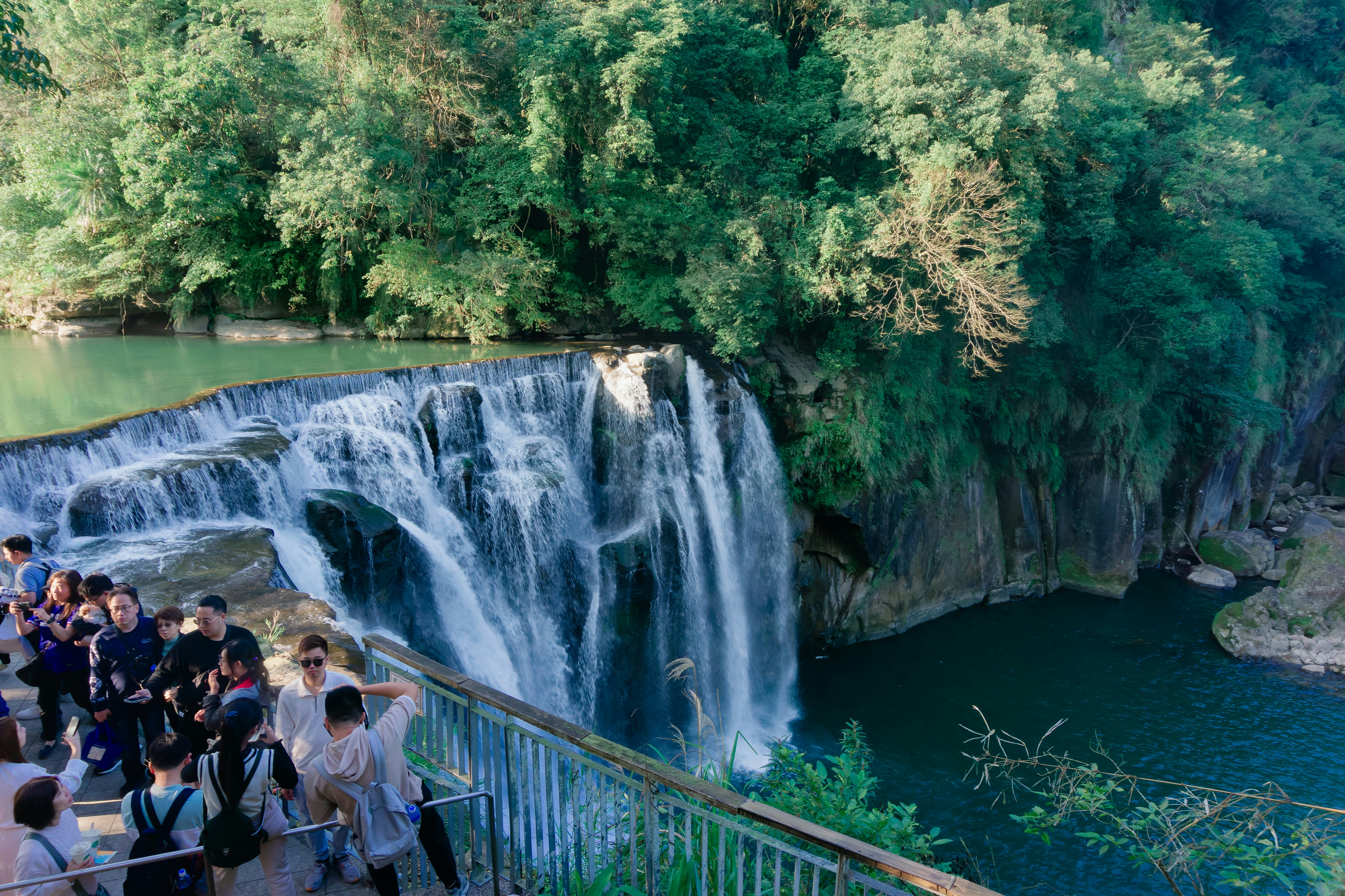 Crowd at a lush riverside overlook beside a cascading waterfall