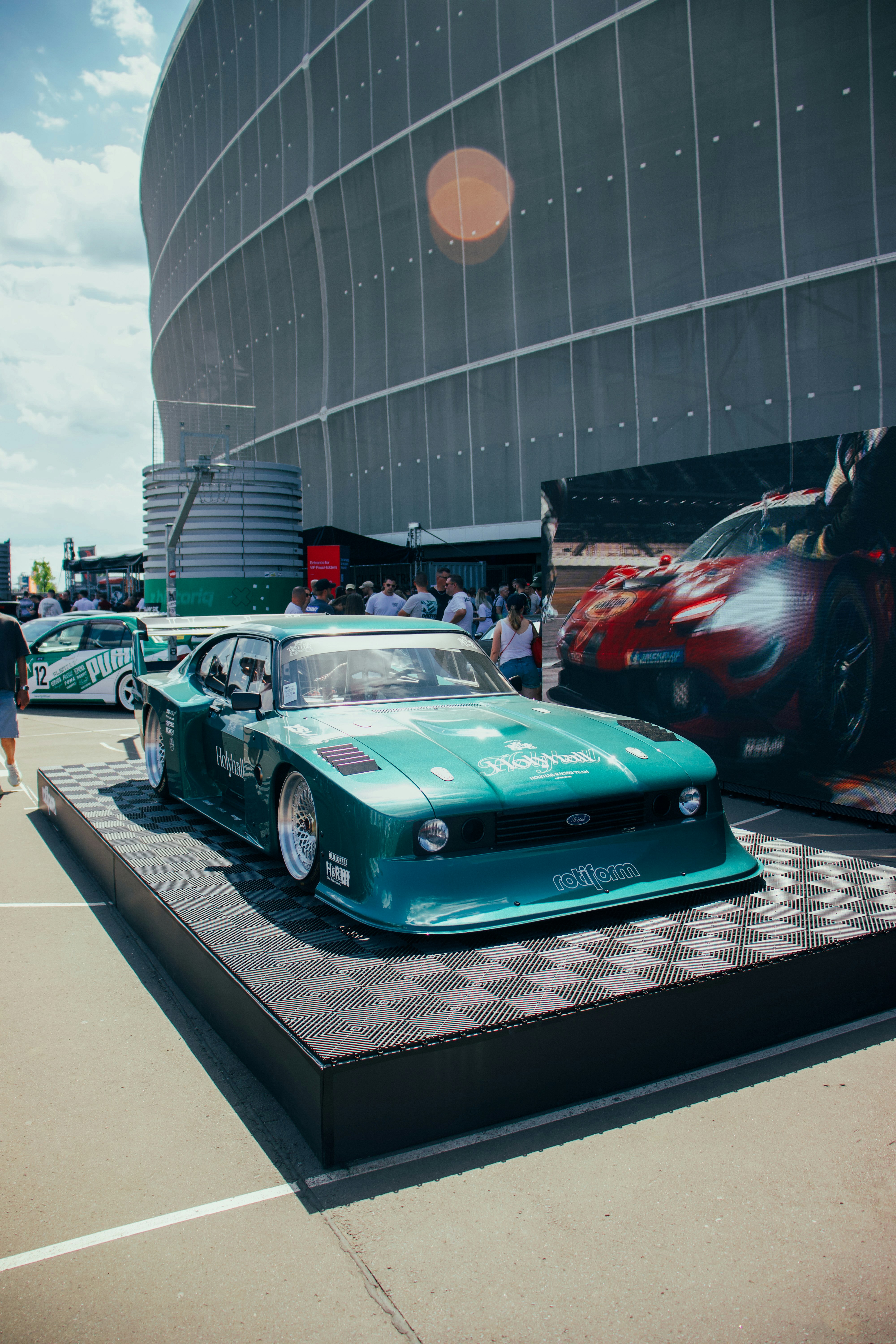 Turquoise racecar on a display platform beneath a curved modern venue
