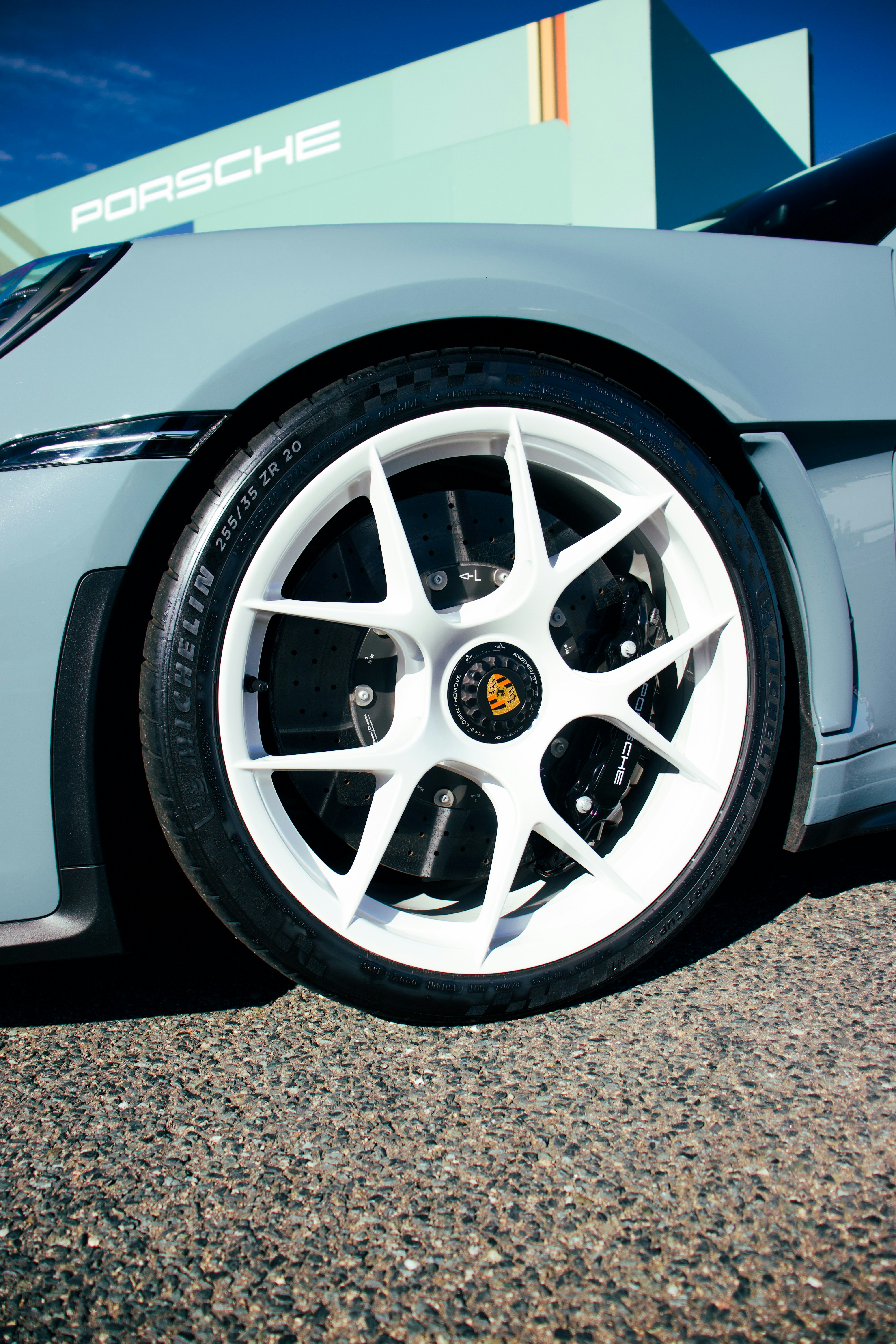 White Porsche wheel under a pale blue body with a Porsche sign in the background