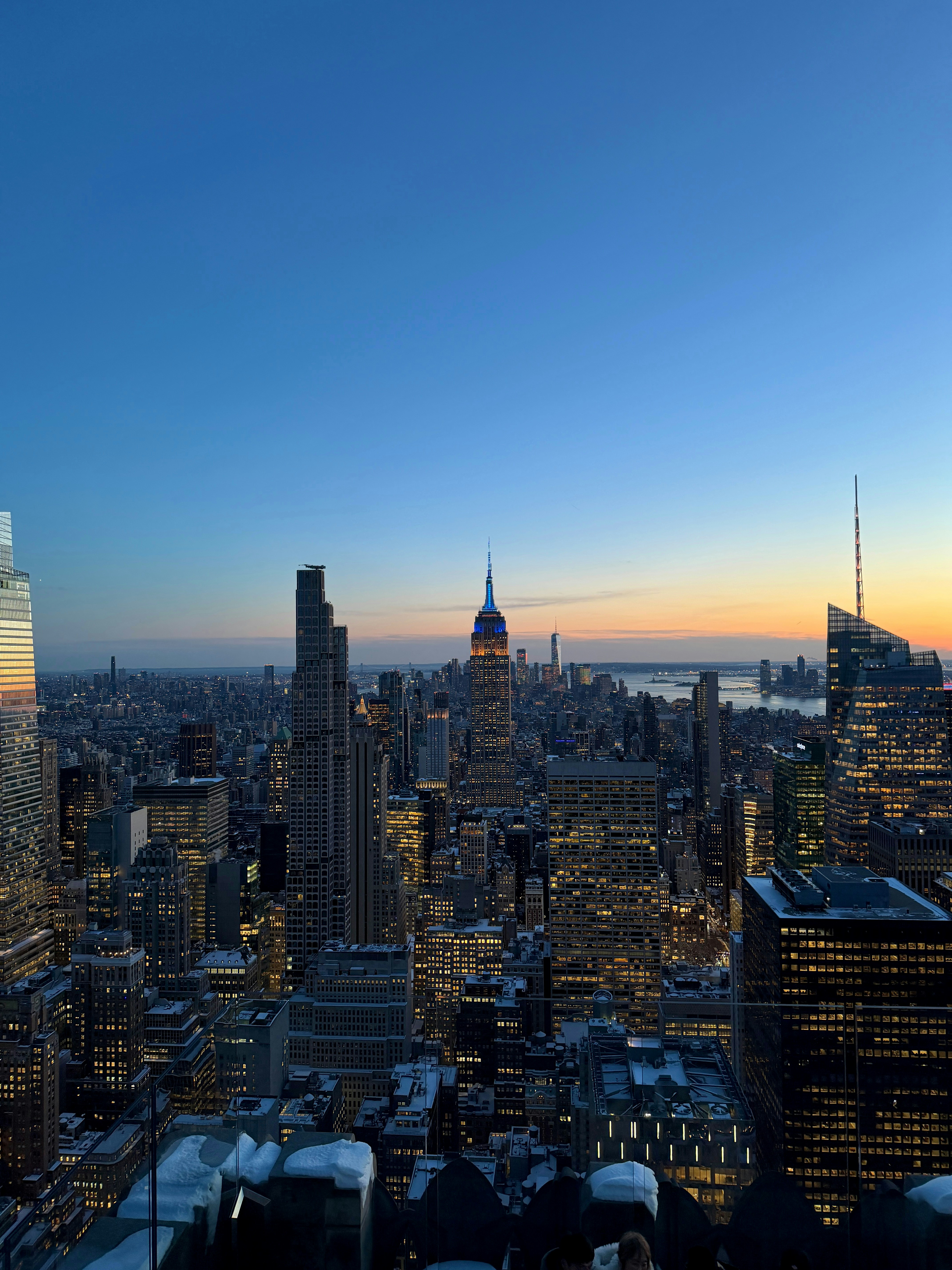 Dense skyscraper skyline at dusk with a warm horizon glow; foreground rooftops lead to a distant vantage point over the water, emphasizing vertical geometry of the cityscape. This is a photographic capture of a cityscape, not a painting or logo.