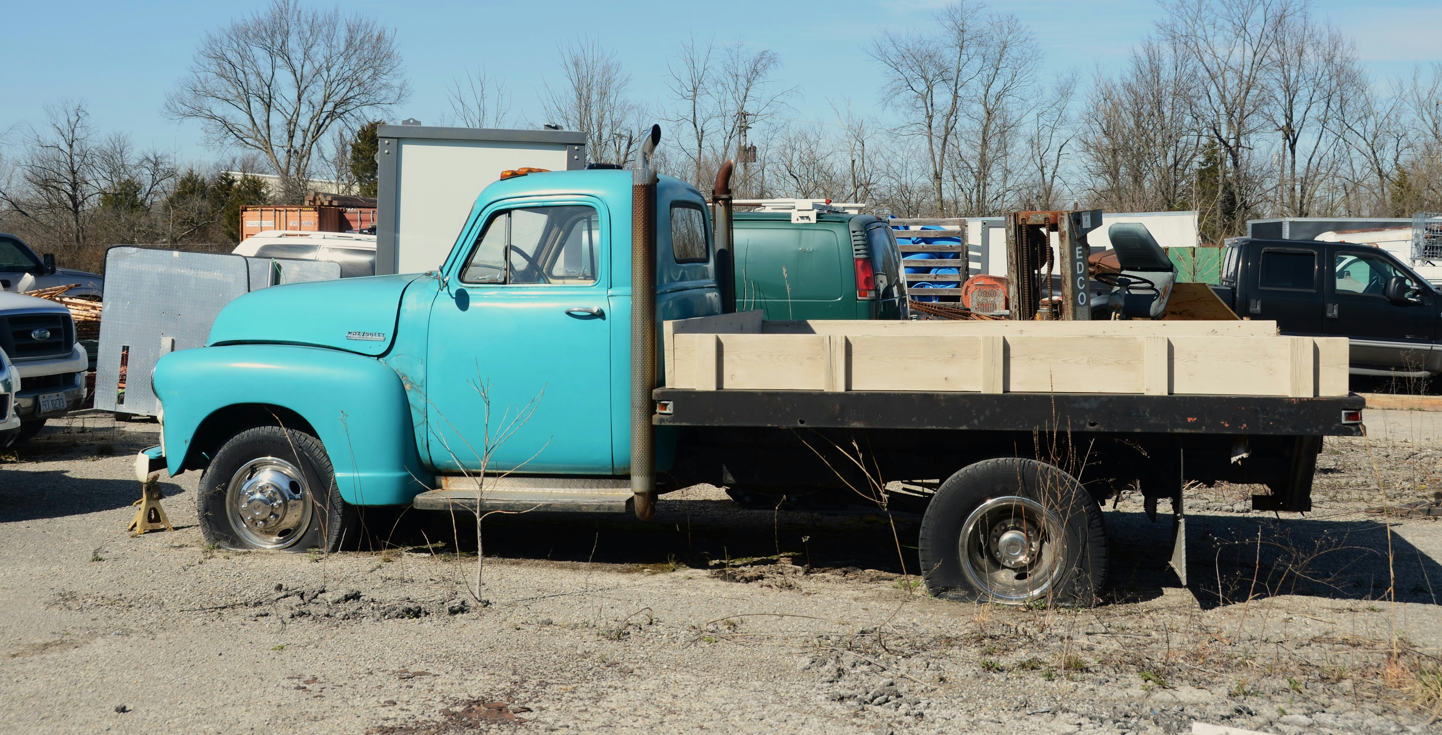 A Weathered teal pickup sits long-quiet among a junkyard of tools and pallets
