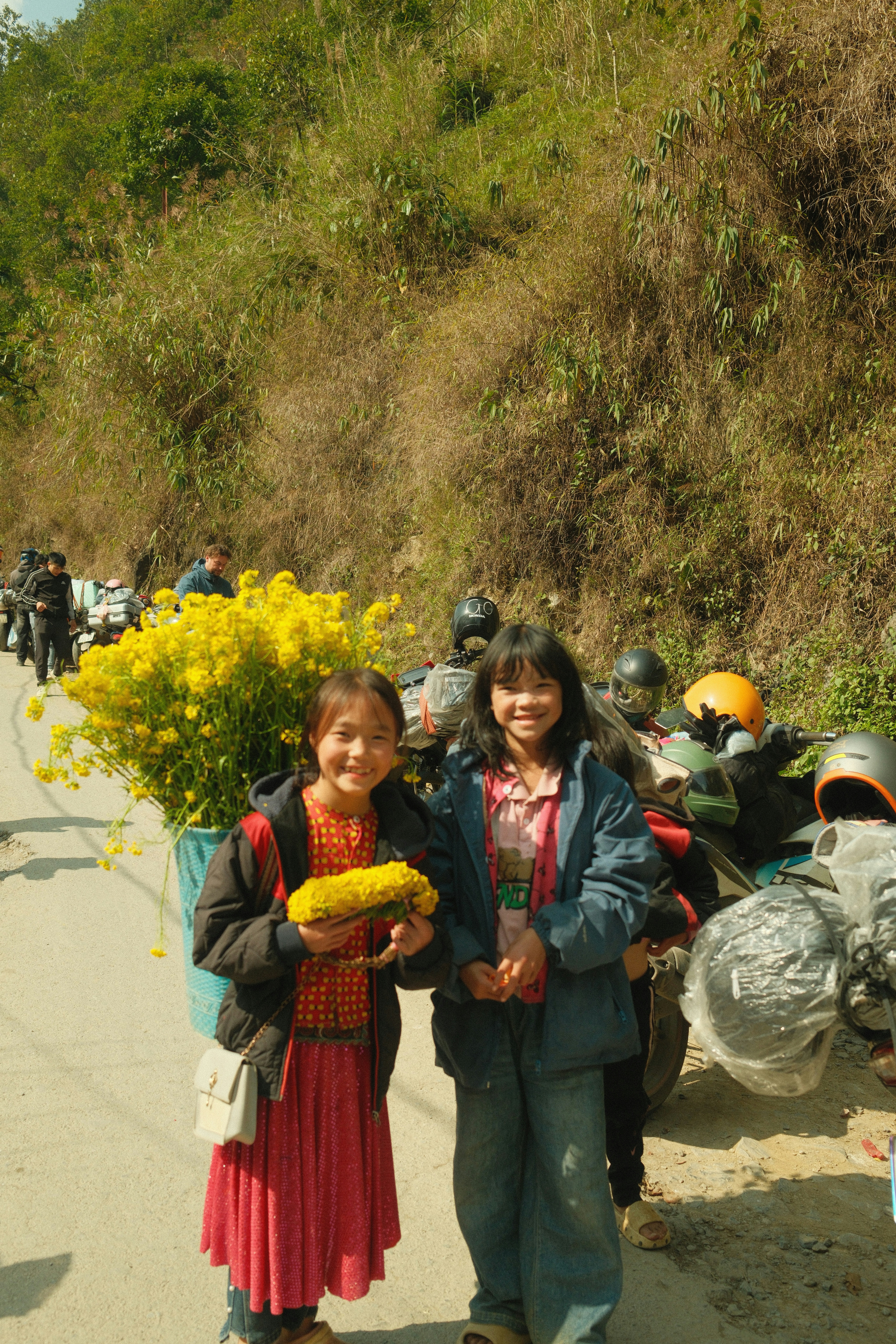 Two friends in a hillside village hold bright yellow flowers amid a line of motorbikes