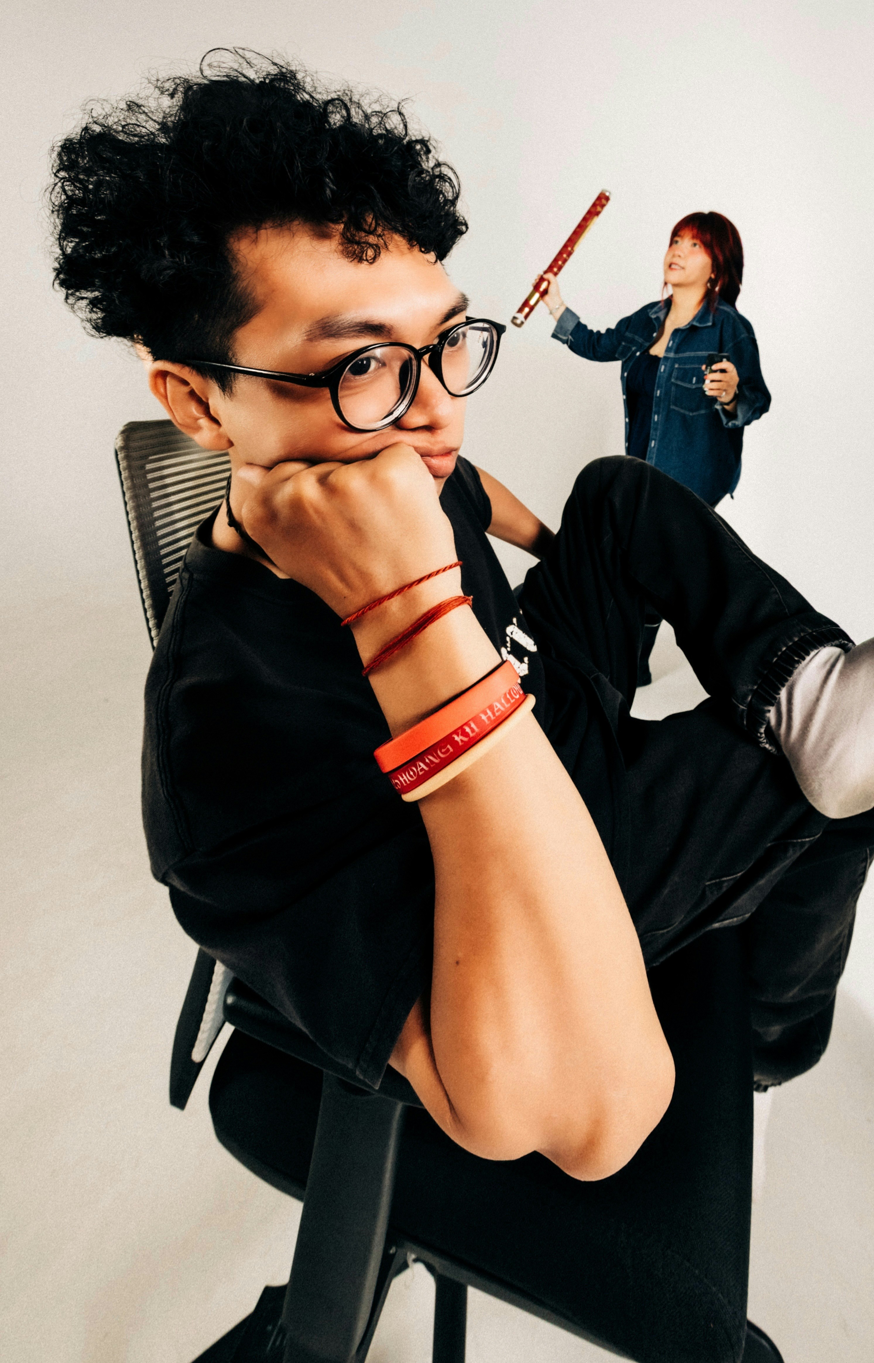 Two figures in a studio scene, one foreground subject seated with a thoughtful pose and a red-braceleted arm, and a guitarist-like figure in the background. The setting is clean with a neutral backdrop, emphasizing the subjects.