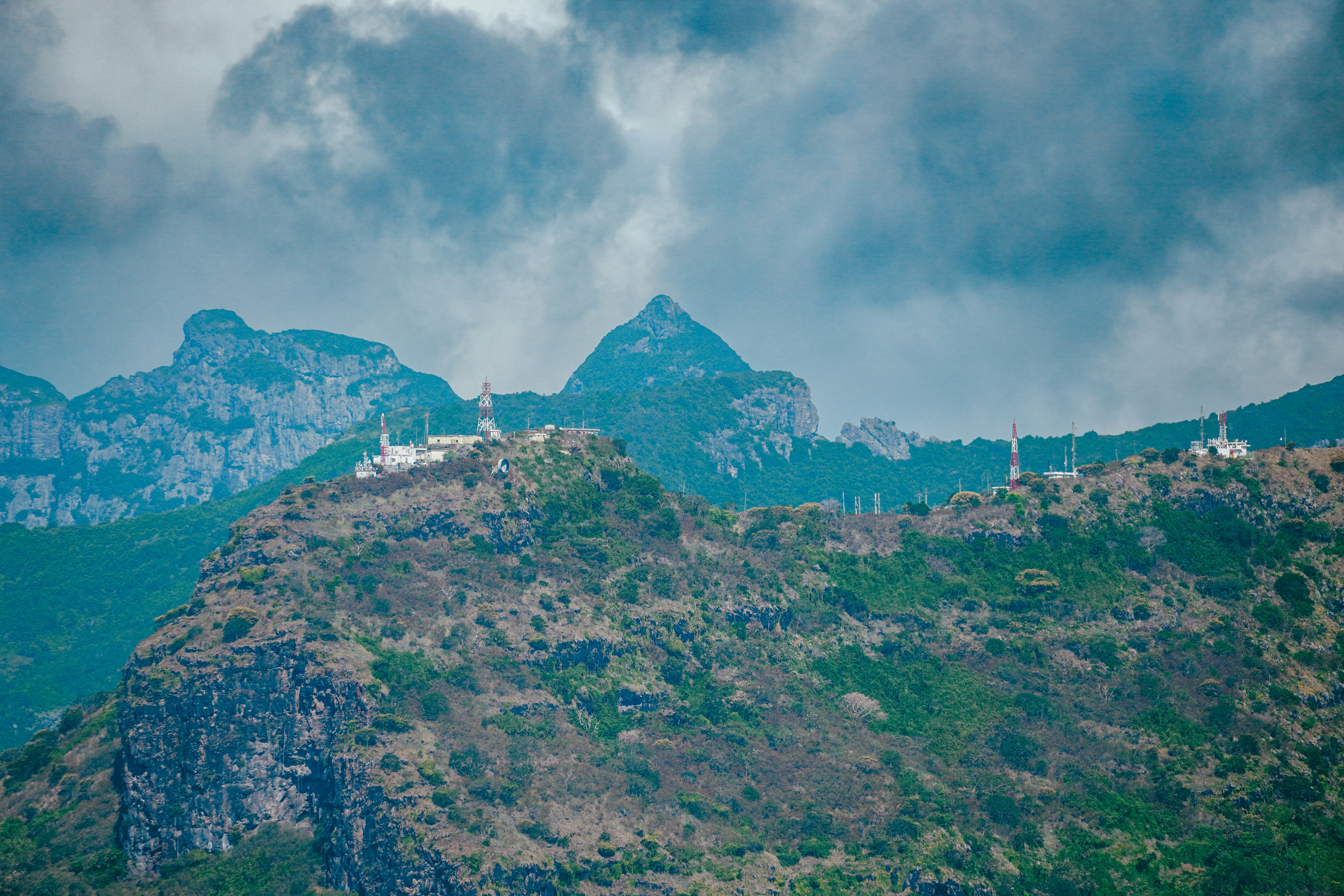 Wind-swept ridge with a line of small structures and antennas on a green-brown hill under a moody, cloud-filled sky.