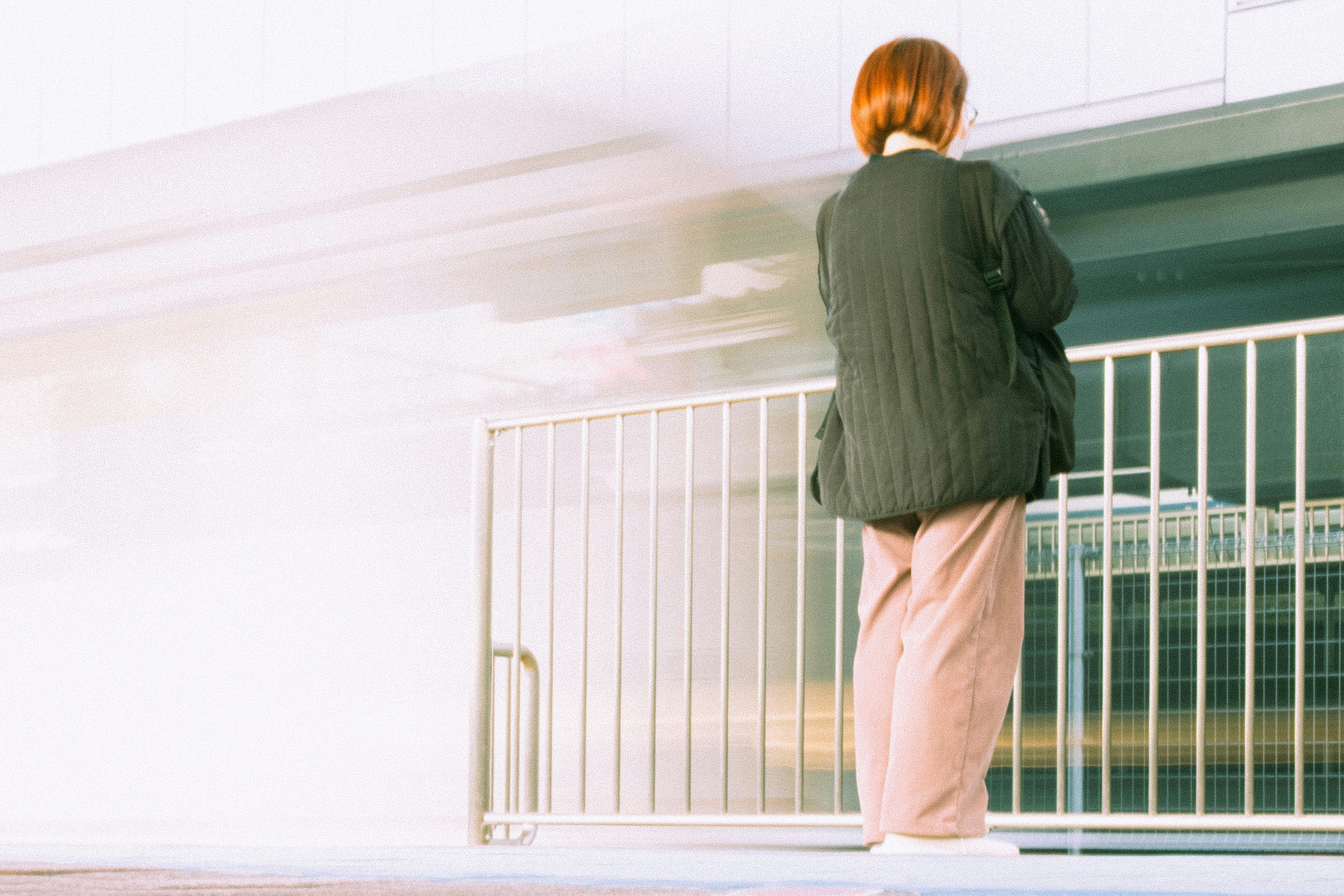 Person in a dark jacket at a railing, walking away on a sunny elevated platform