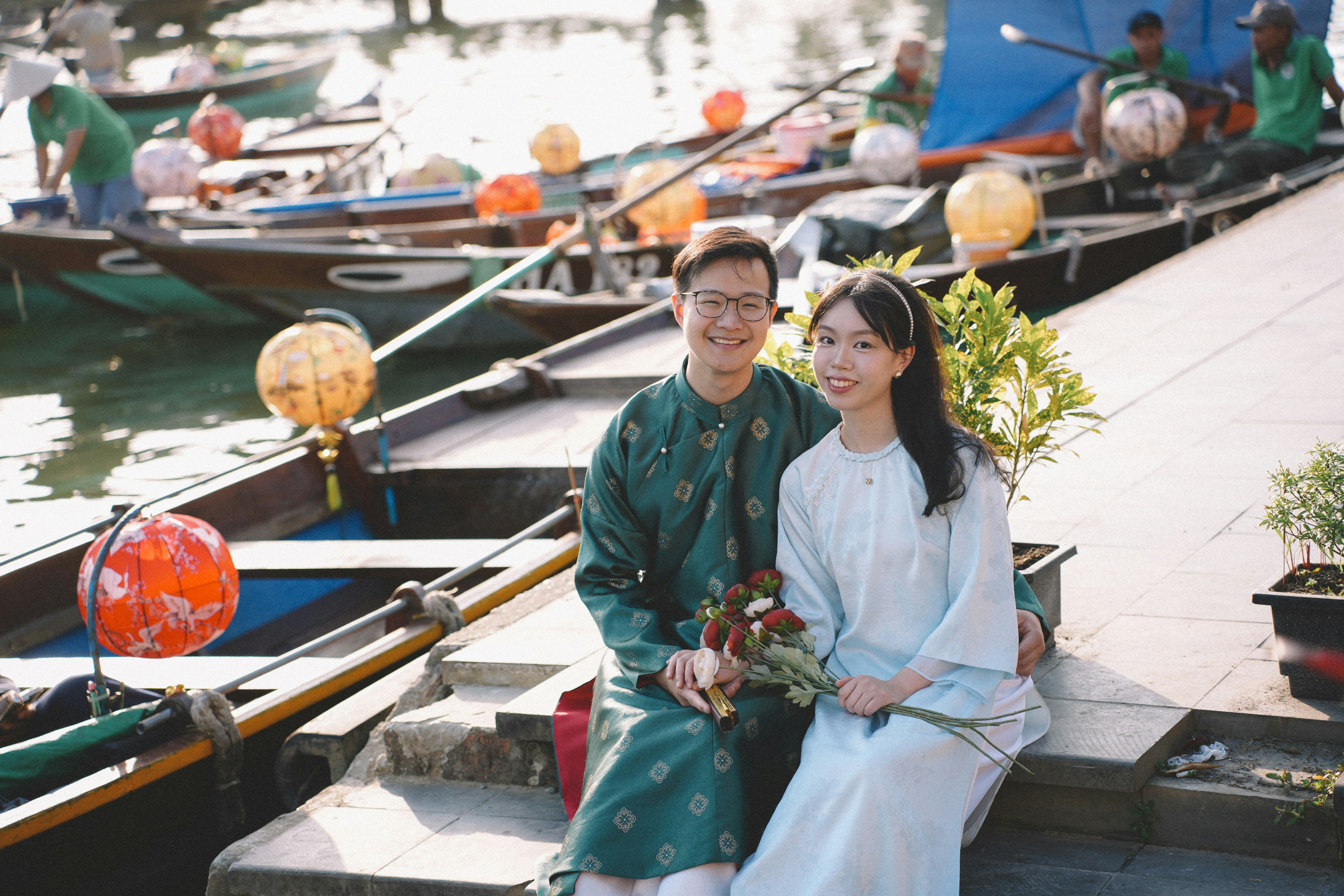 Couple in traditional attire by the canal, holding flowers at a sunlit riverside